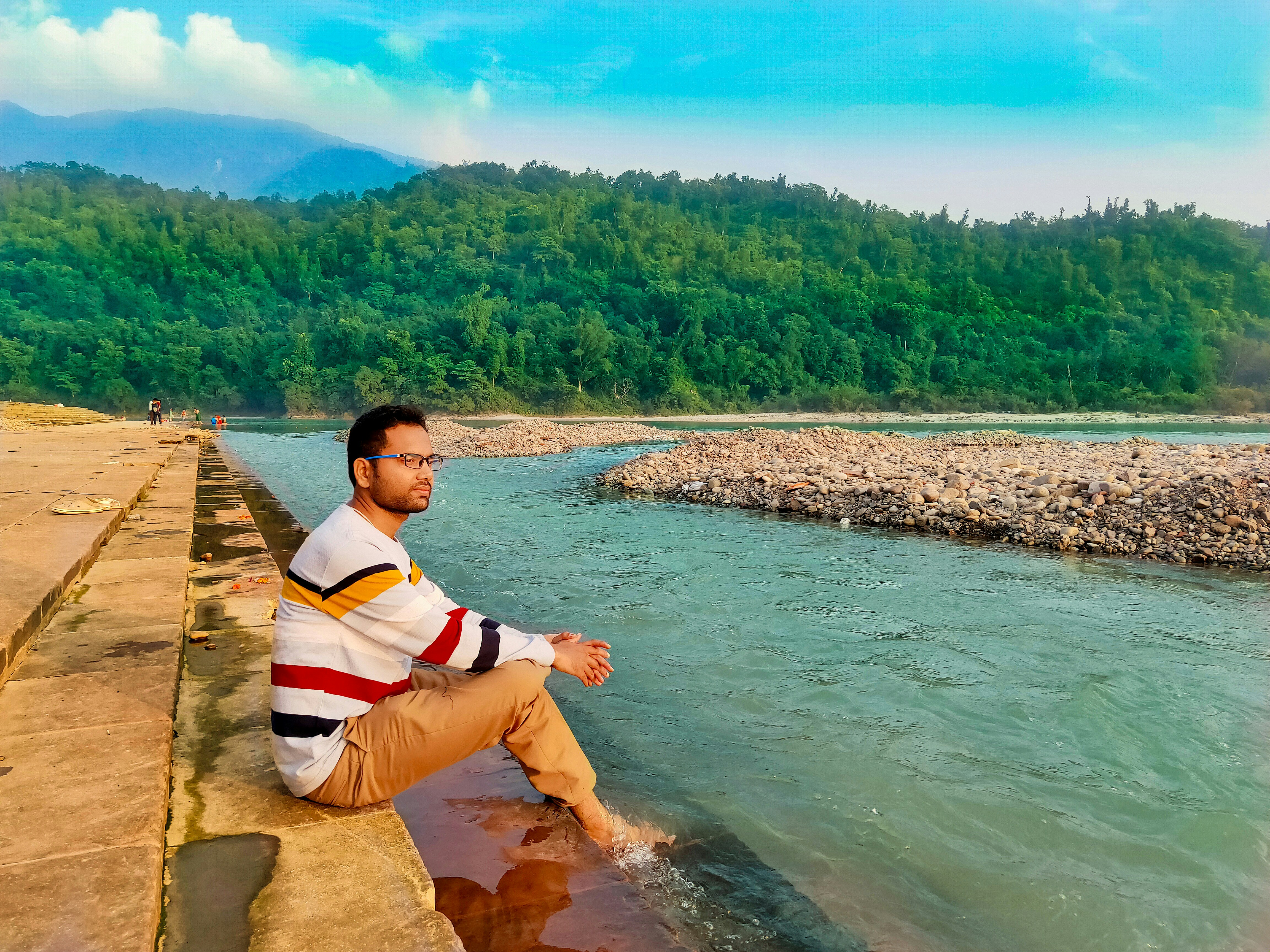 Man sits on a concrete jetty beside a turquoise river, with forested hills in the distance, capturing a quiet, contemplative moment.