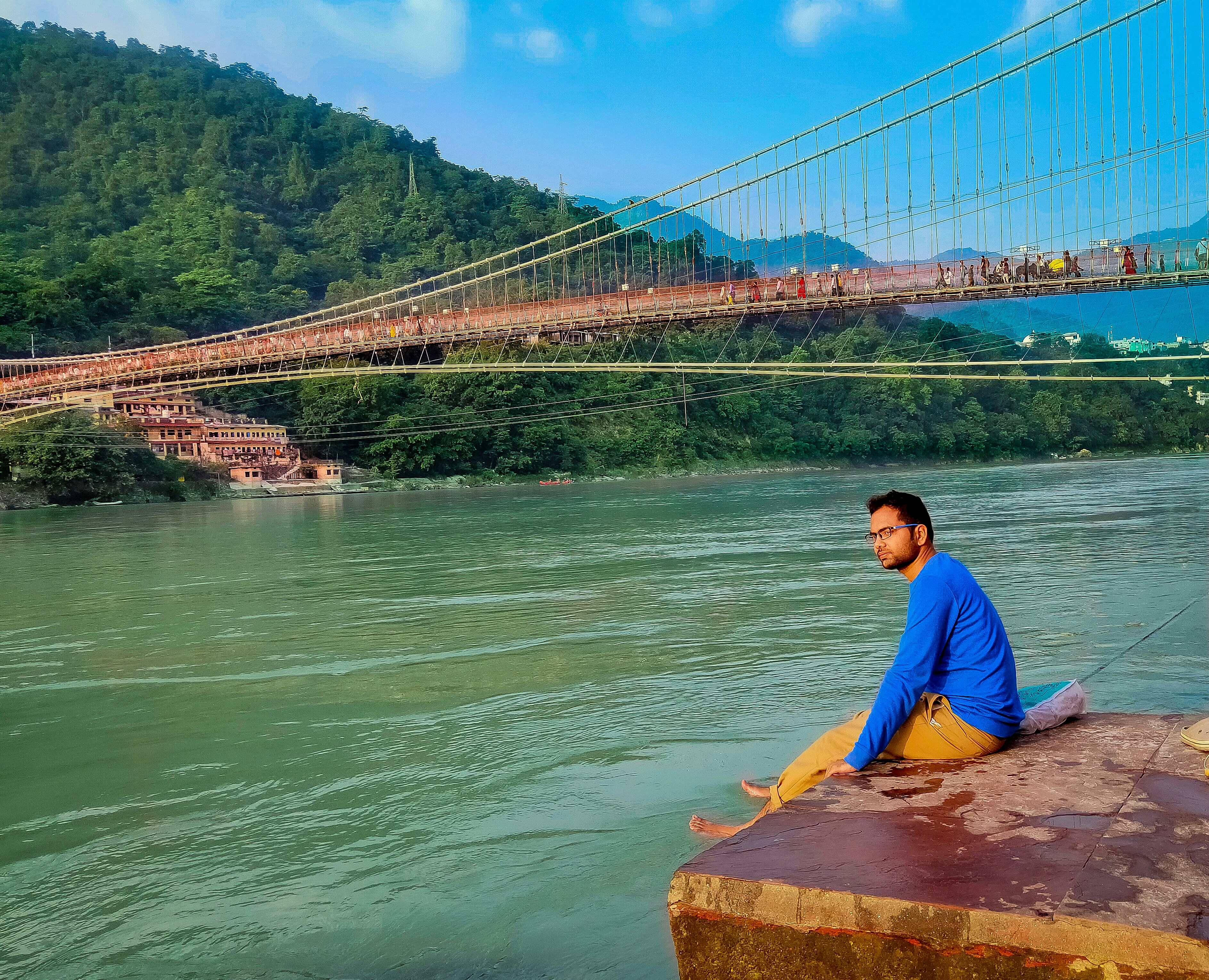 Person in blue sitting on a rock by the river with a suspension bridge in the background.