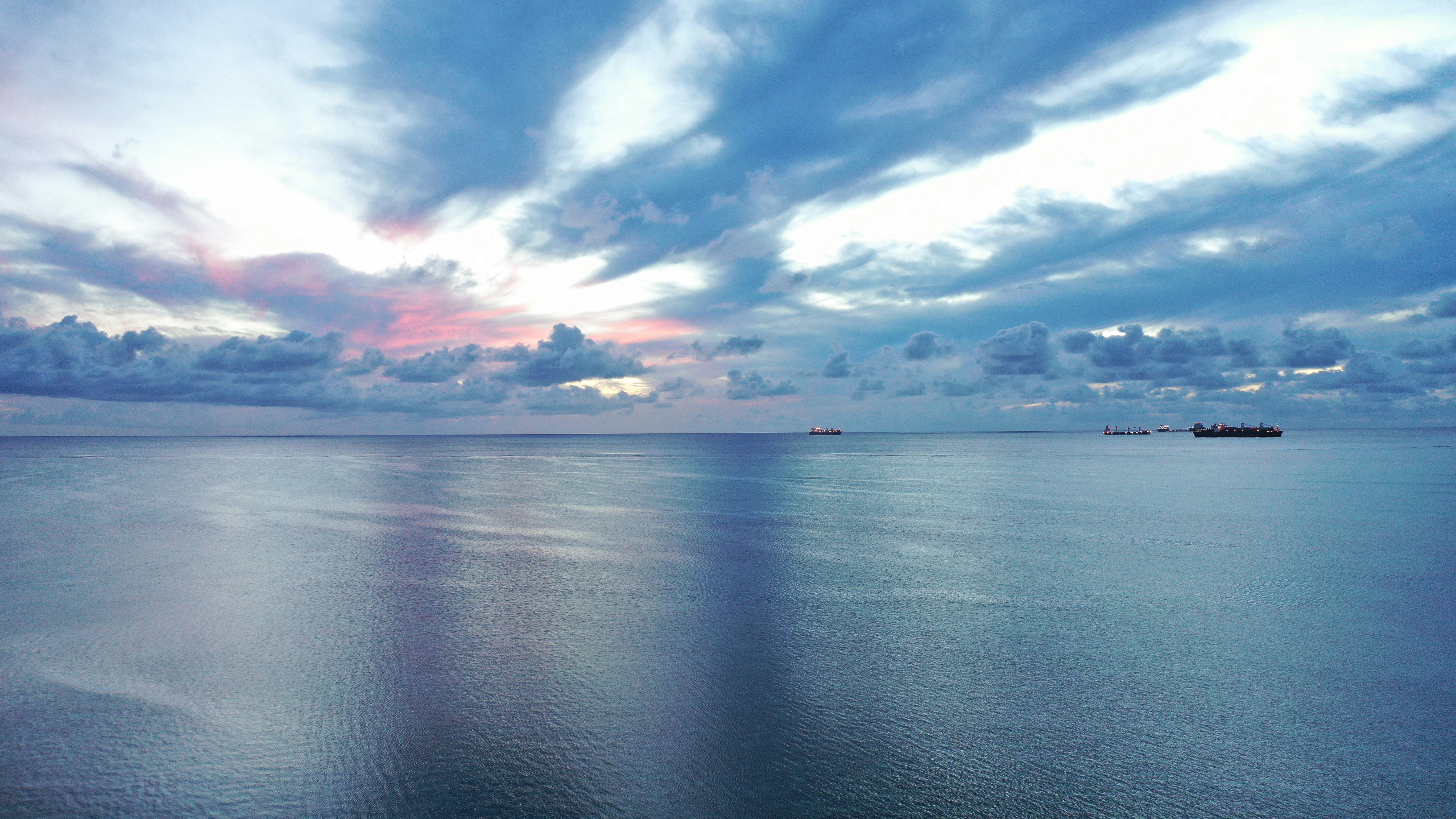 Vast ocean under a dramatic sky, featuring soft hues of pink and blue as the sun sets. Cargo ships dot the horizon, adding depth to the serene seascape.