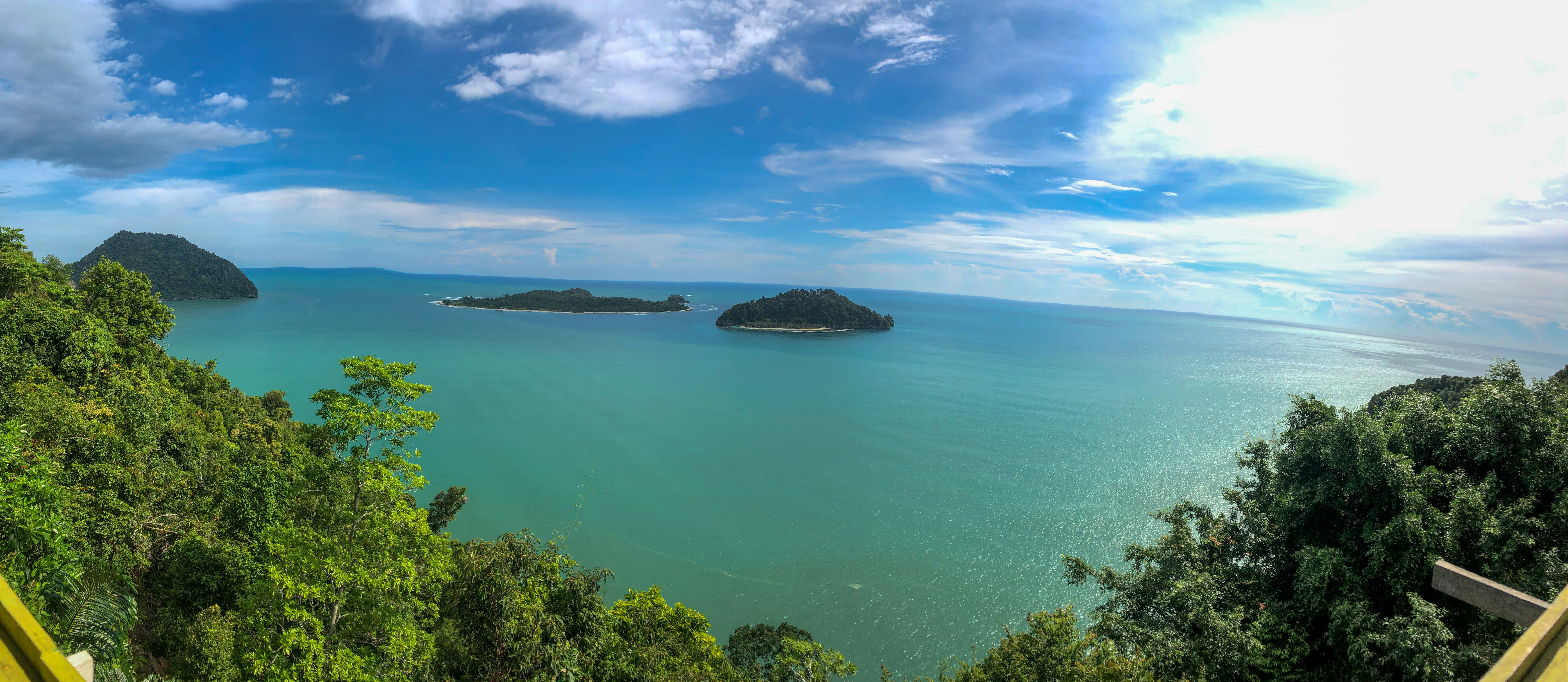 Vibrant turquoise waters stretch towards distant islands under a vast blue sky, framed by lush greenery. A panoramic view captures the tranquil beauty of the coastal landscape.