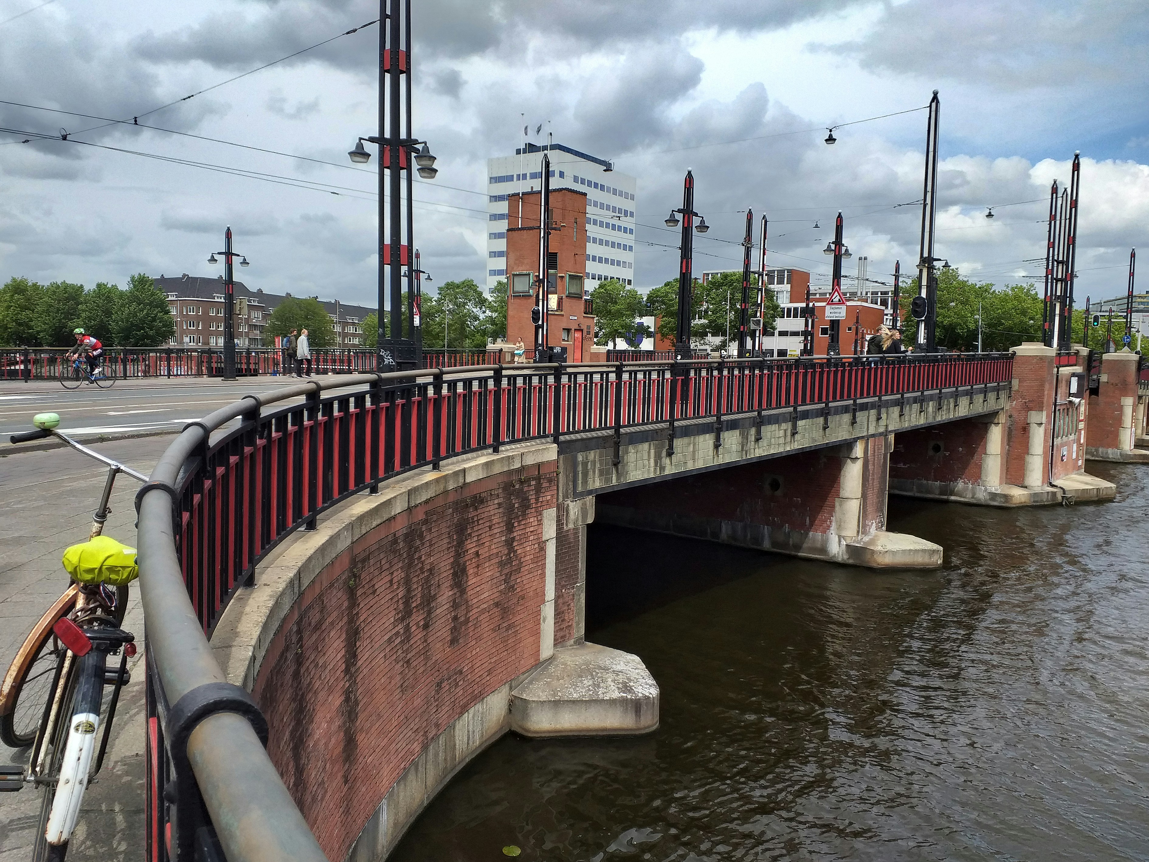 A bicycle rests against a curved railing on a red-accented bridge overlooking a calm river, framed by modern architecture under a cloudy sky.