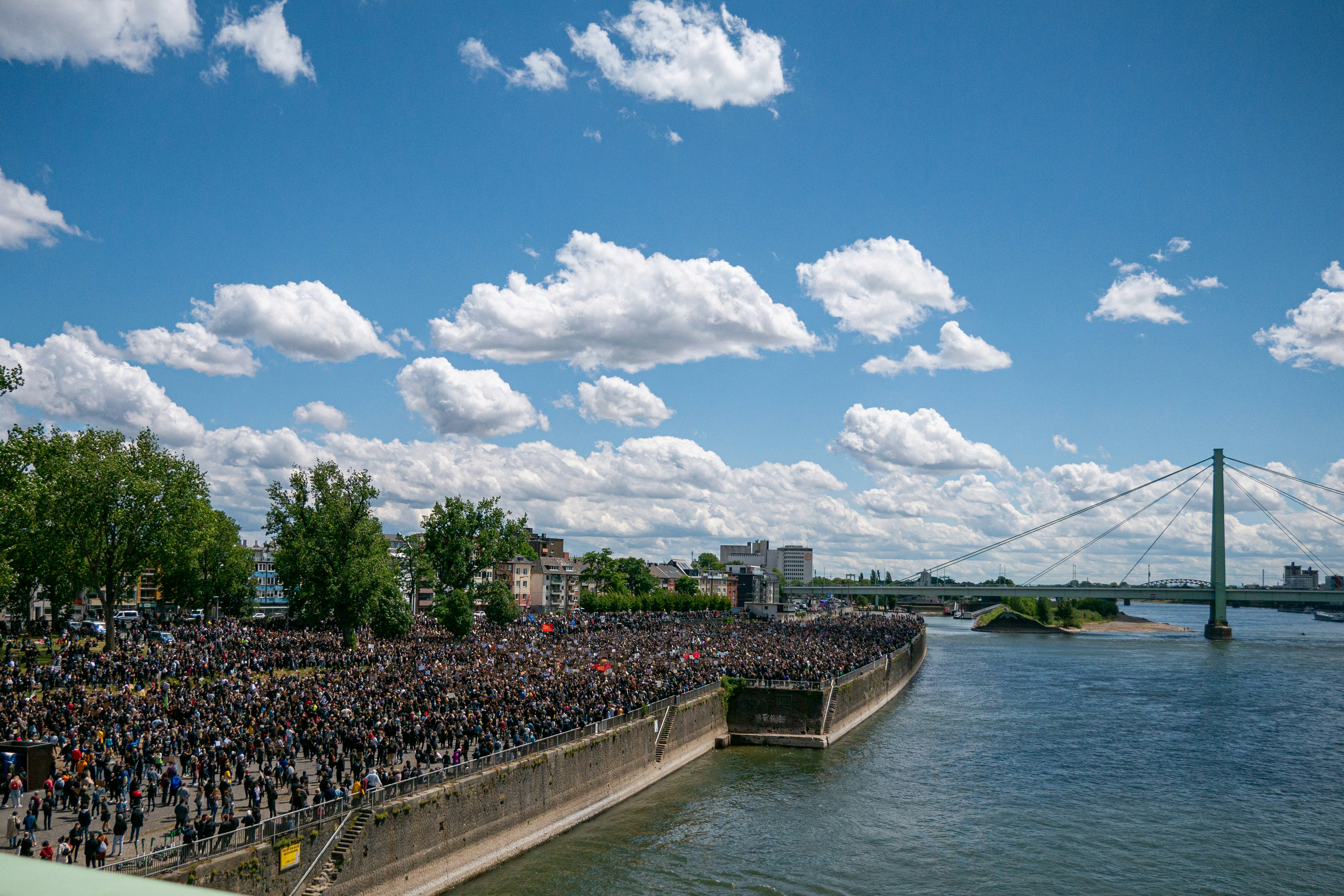 Crowd gathered along a riverside promenade beneath a vibrant blue sky with scattered clouds.