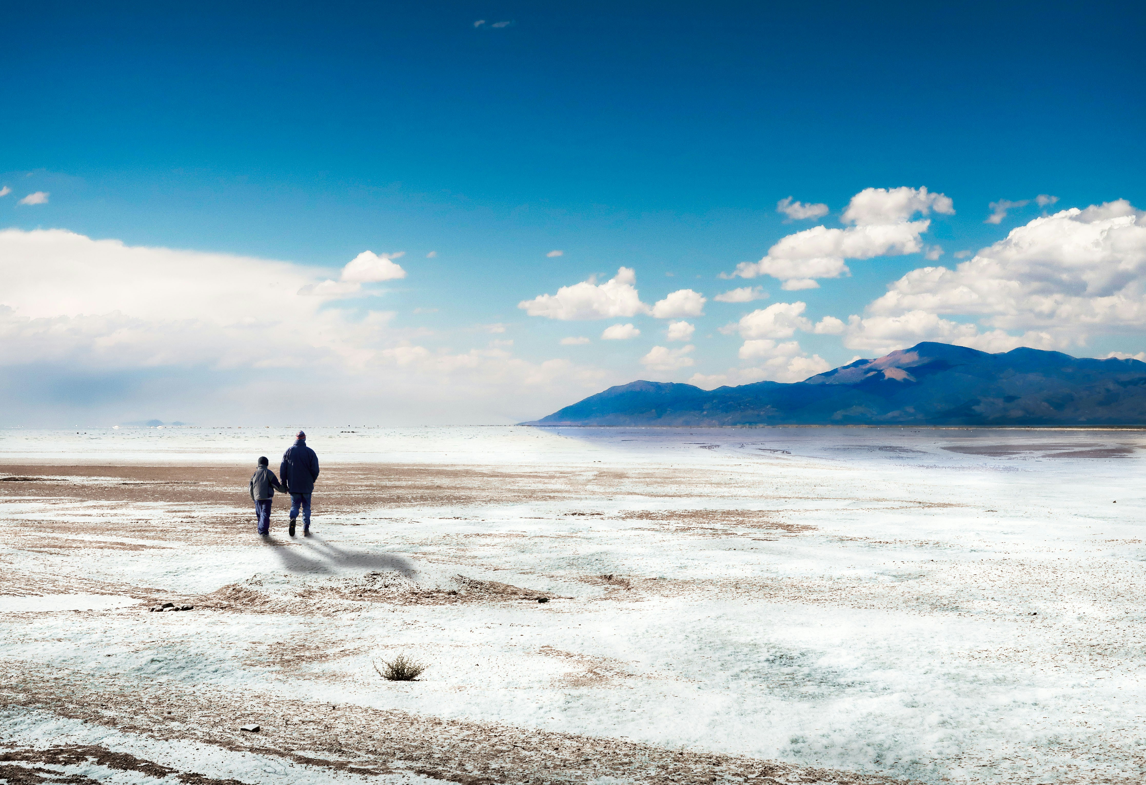Two figures walk across a vast, bright salt flat with mountains in the distance under a clear blue sky.
