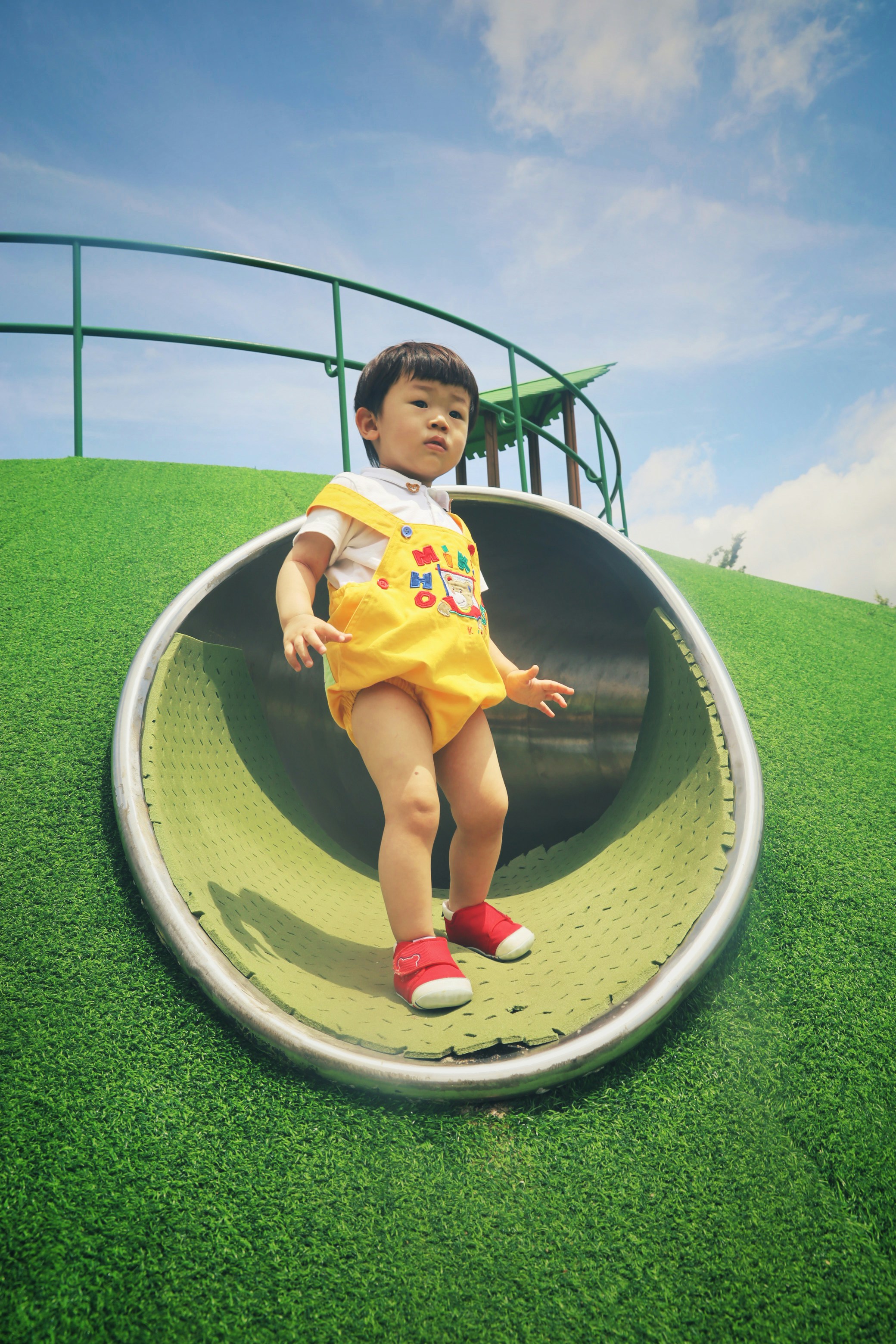 Child in yellow overalls playing on a slide in a vibrant playground, surrounded by green turf and bright blue skies.
