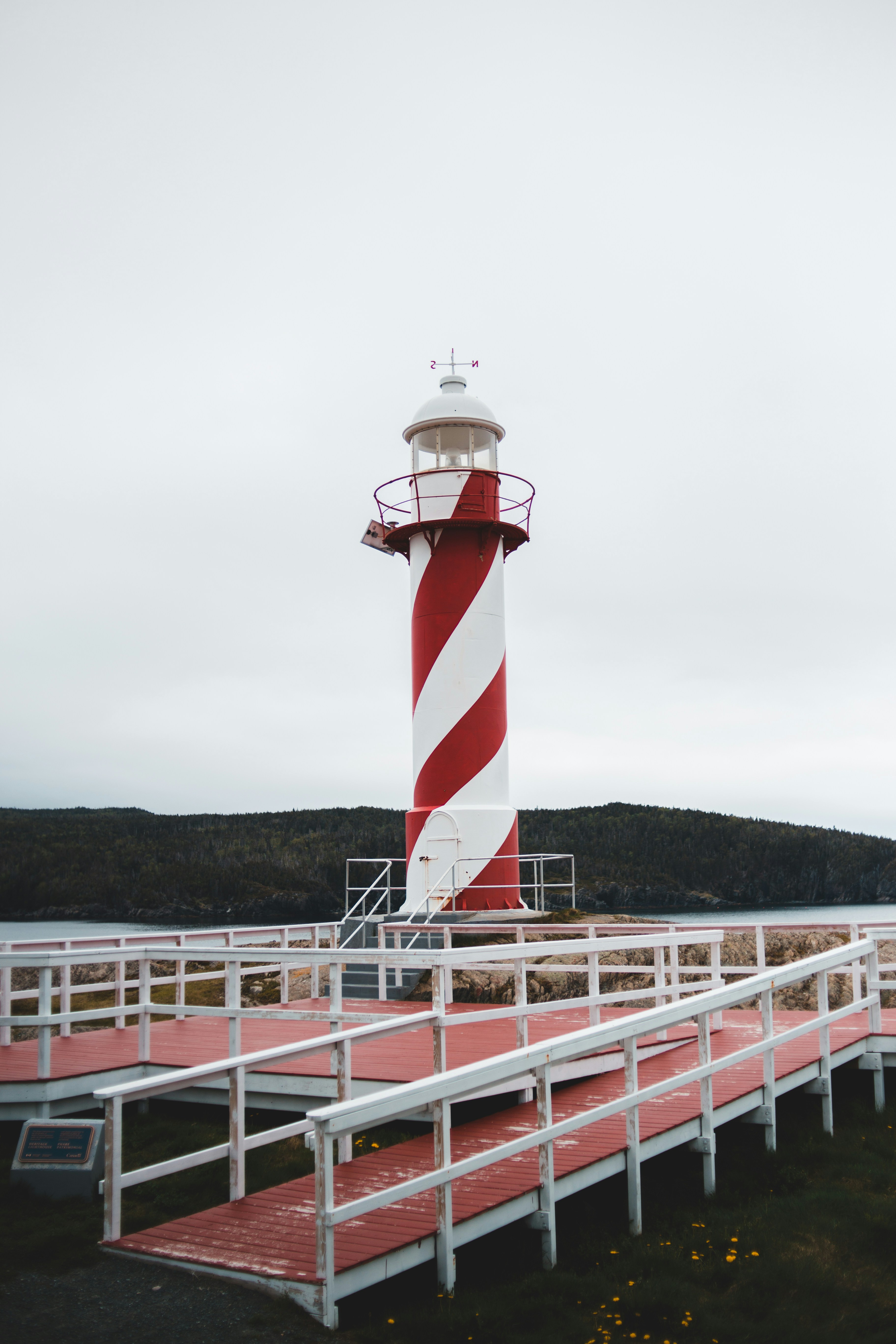 A vibrant red and white striped lighthouse stands tall against a cloudy sky, surrounded by a wooden walkway. The structure serves as a beacon in a serene coastal landscape.