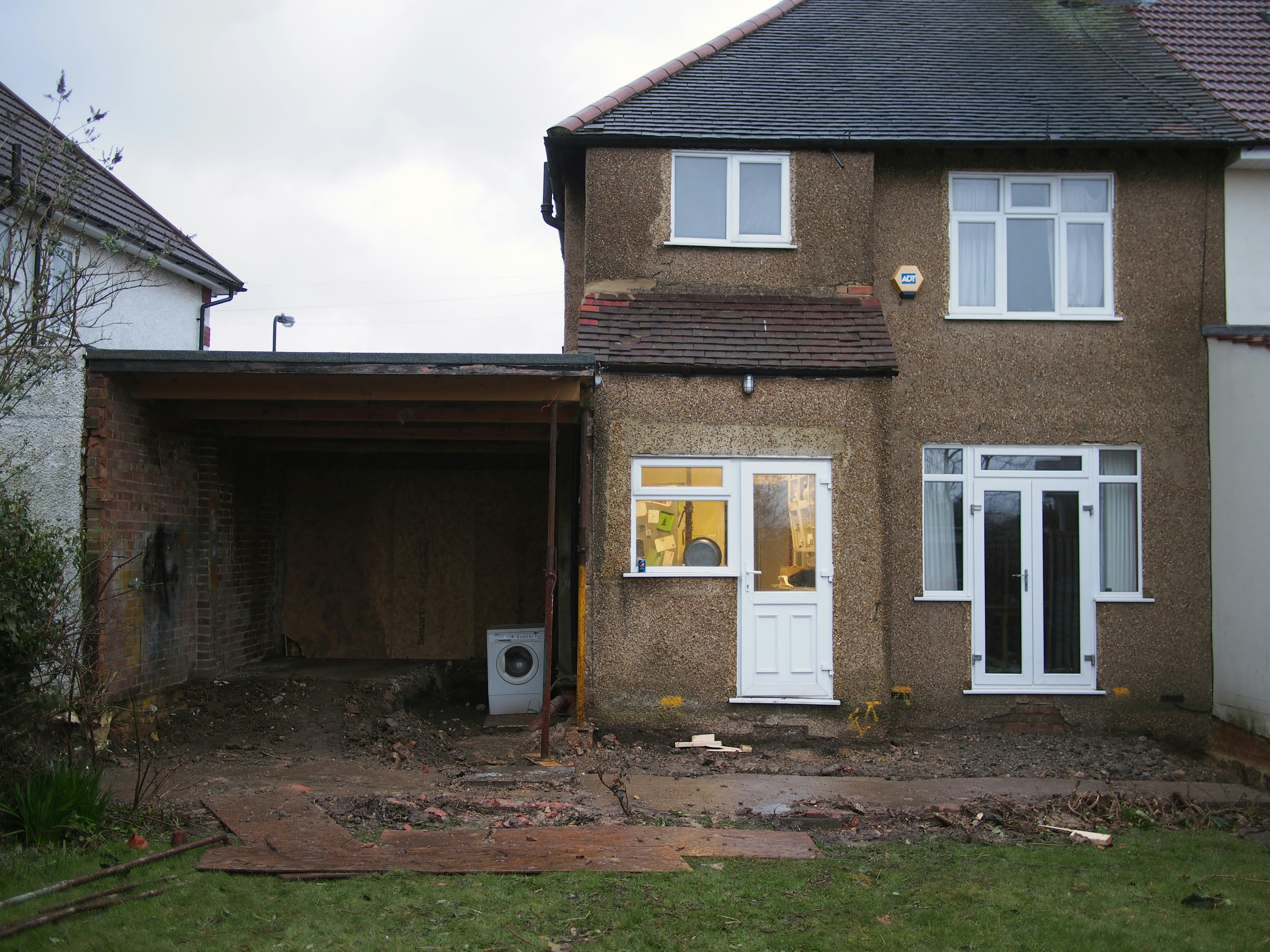 A semi-detached house undergoing renovation, featuring a partially constructed extension and visible construction materials. The scene captures the essence of home improvement.
