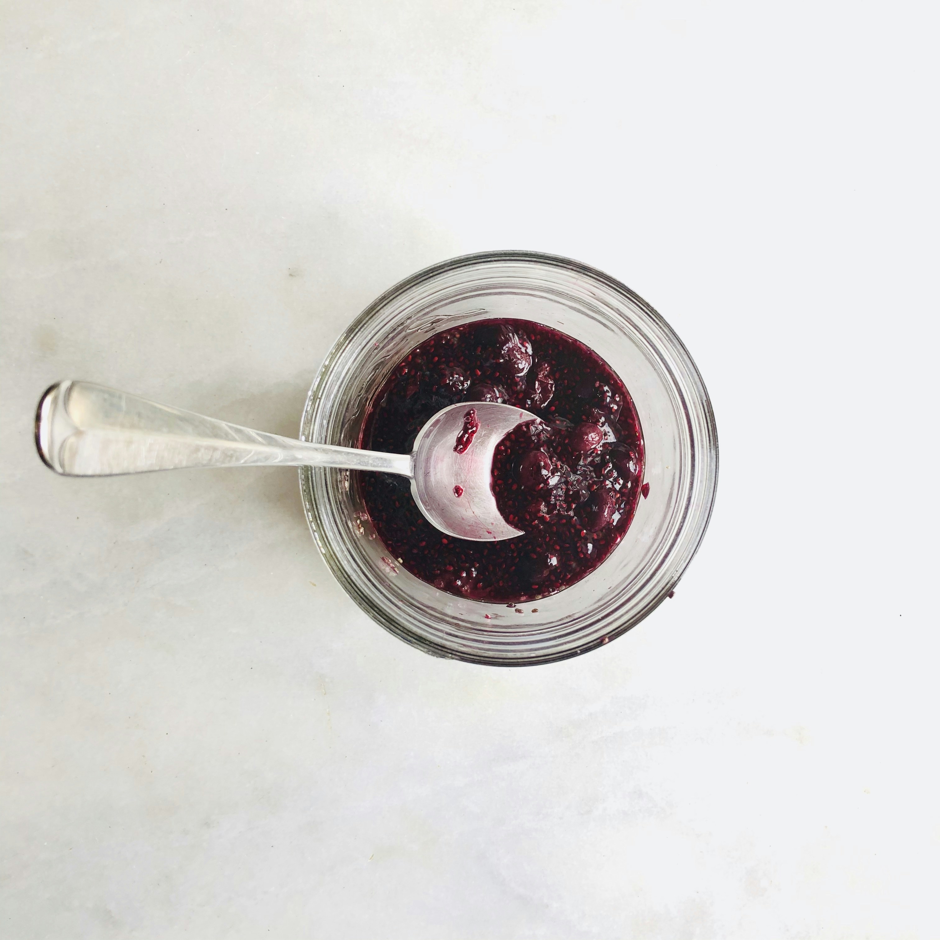 A jar filled with rich, dark berry jam and a silver spoon resting inside, set against a light, neutral background.
