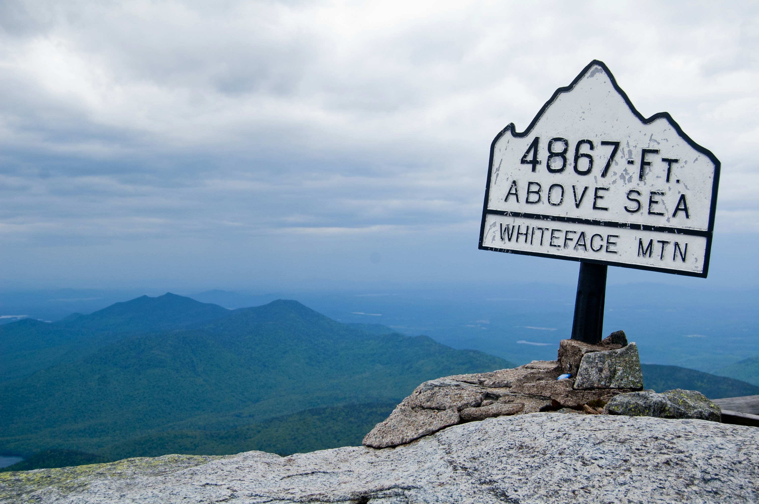 Wooden signage on rocky summit with expansive green mountains under a cloudy sky.