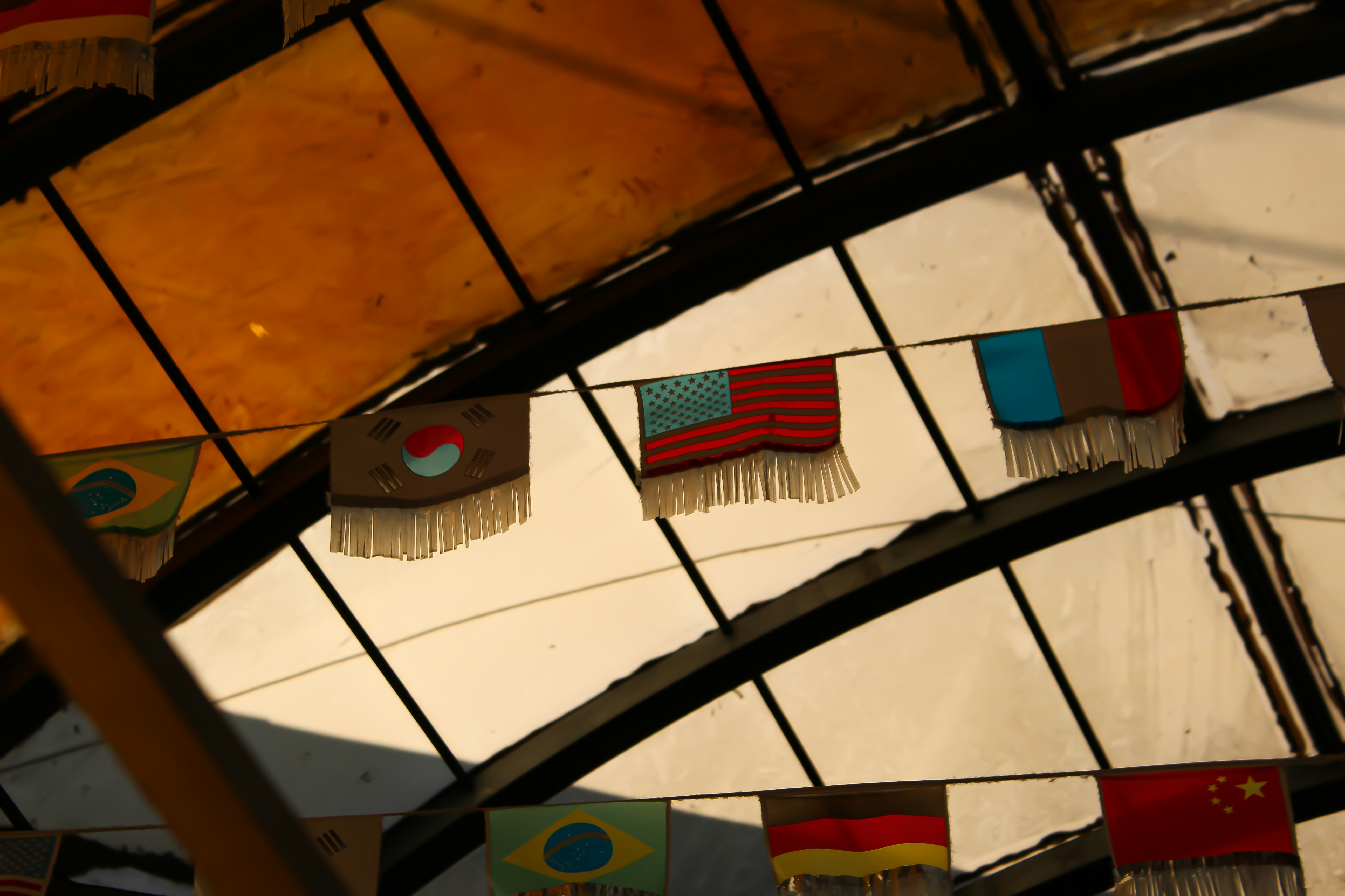 Colorful international flags hanging beneath a translucent, amber-tinted roof, creating a warm and inviting atmosphere.