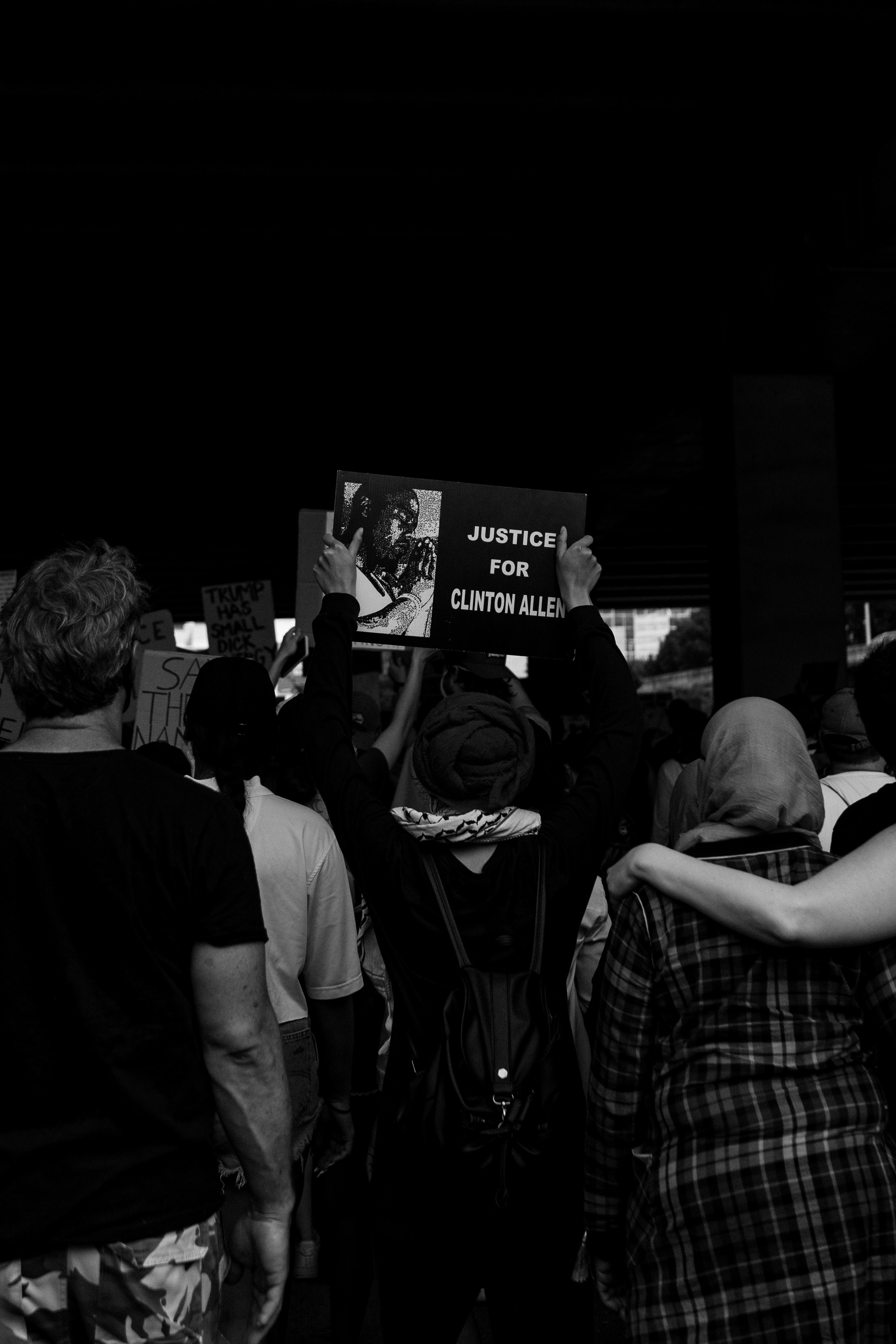 A crowd gathers under an overpass, holding signs in support of Clinton Allen, emphasizing the collective demand for justice.