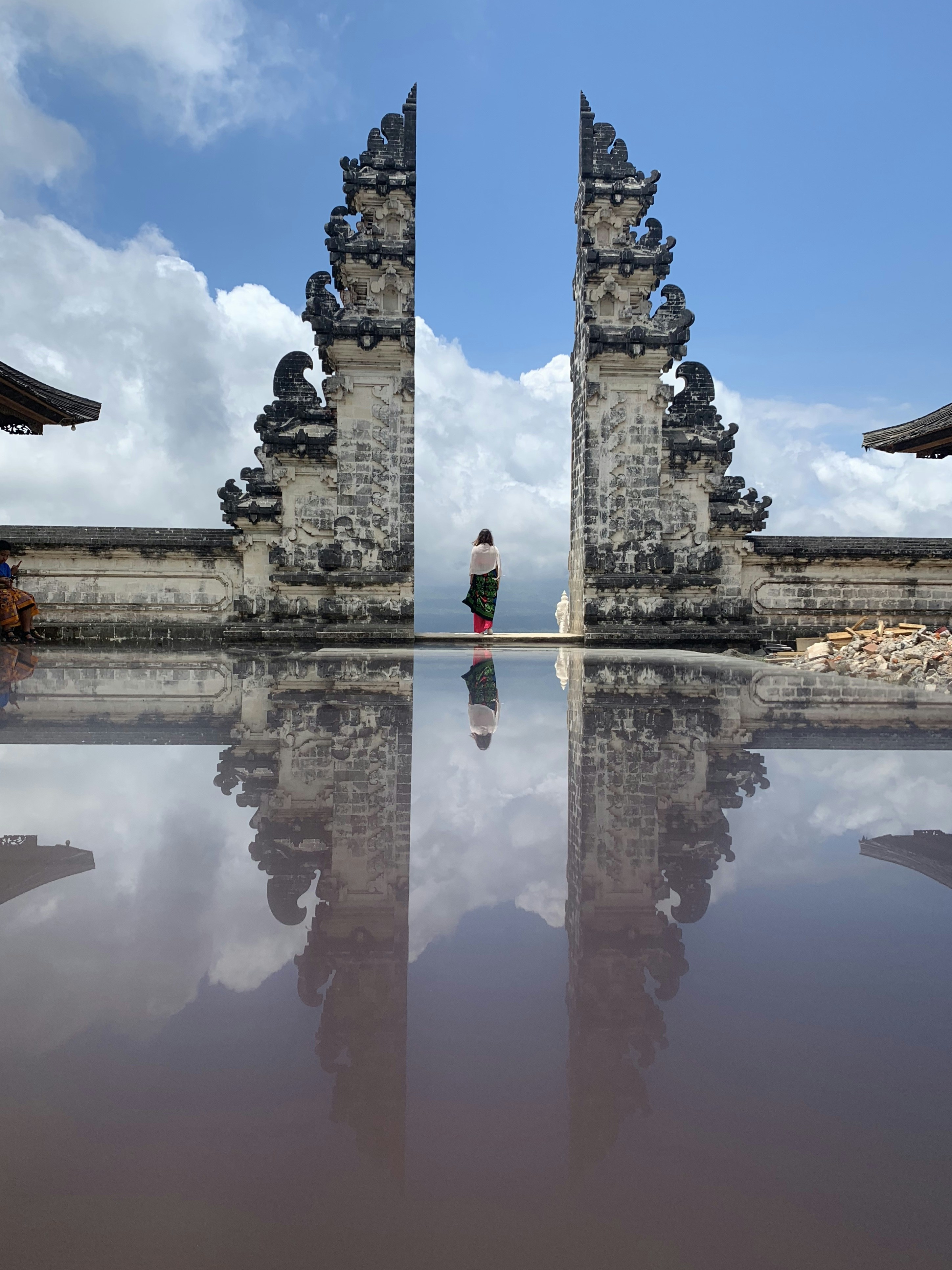 Intricate stone gates reflecting in tranquil water with a figure standing in contemplation. The scene captures a moment of peace and connection with nature.
