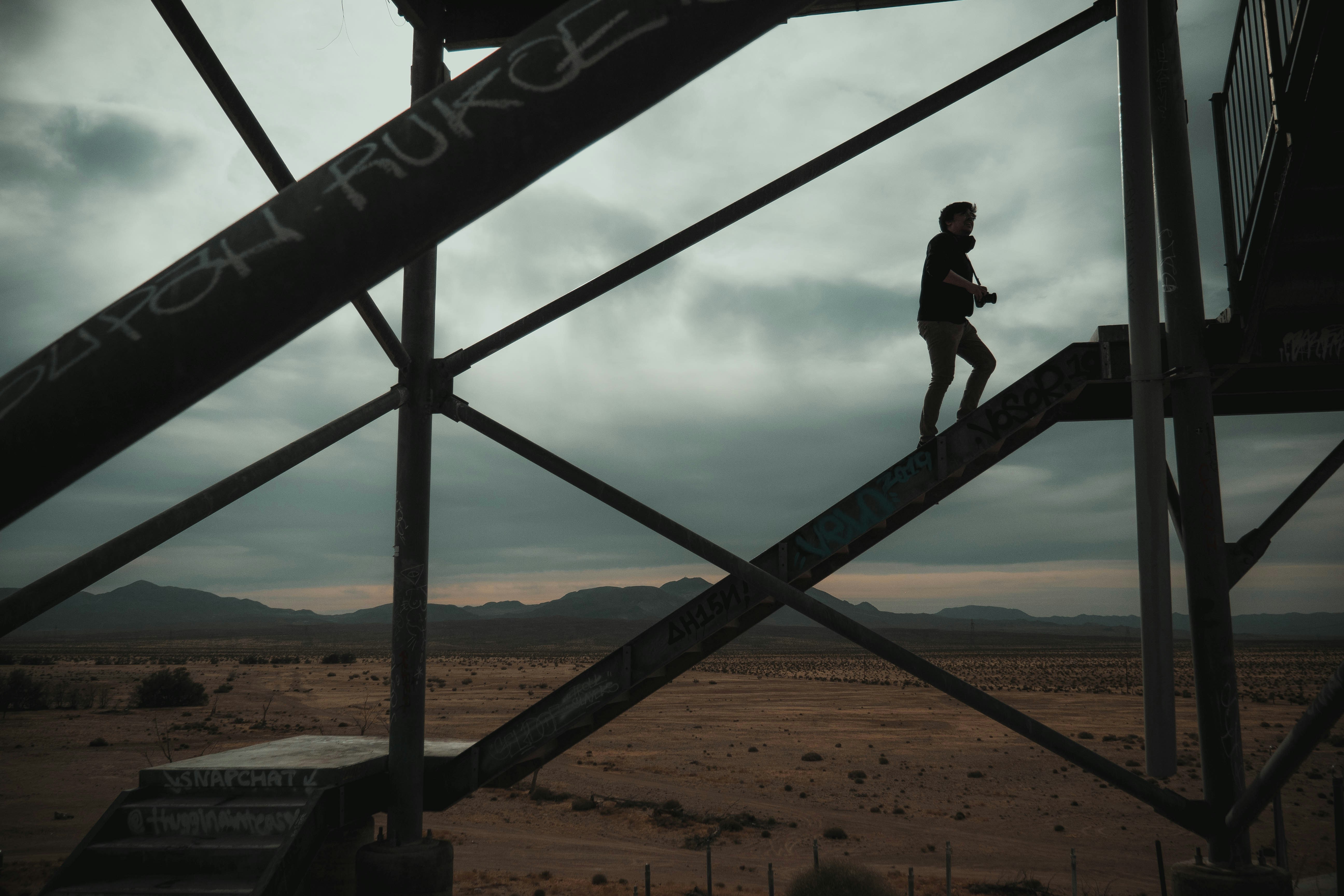 Silhouette of a person ascending a metal staircase in an abandoned waterpark against a barren desert backdrop.