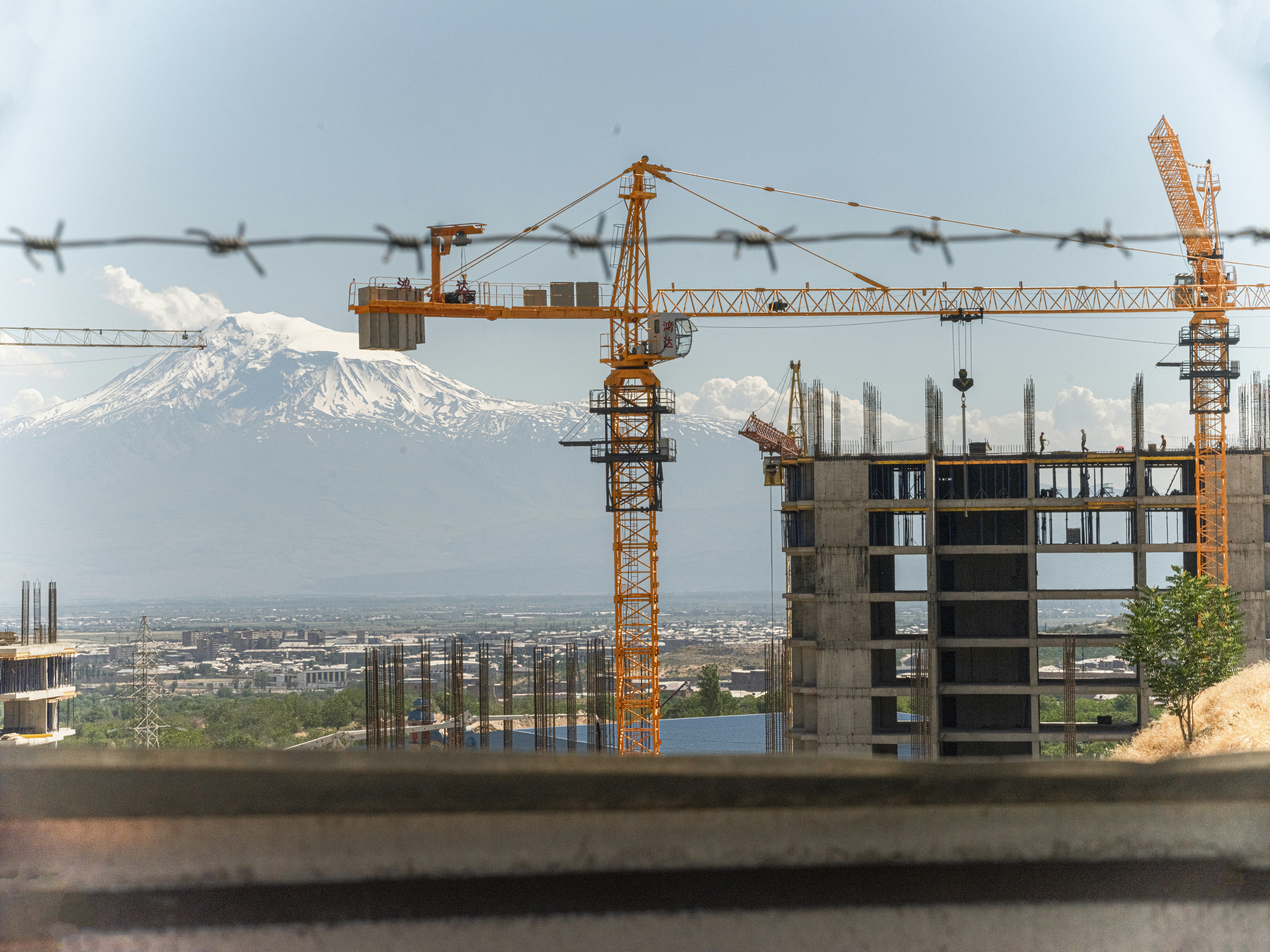 Construction site framed by barbed wire, with a snow-capped mountain in the background, symbolizing the intersection of progress and nature.