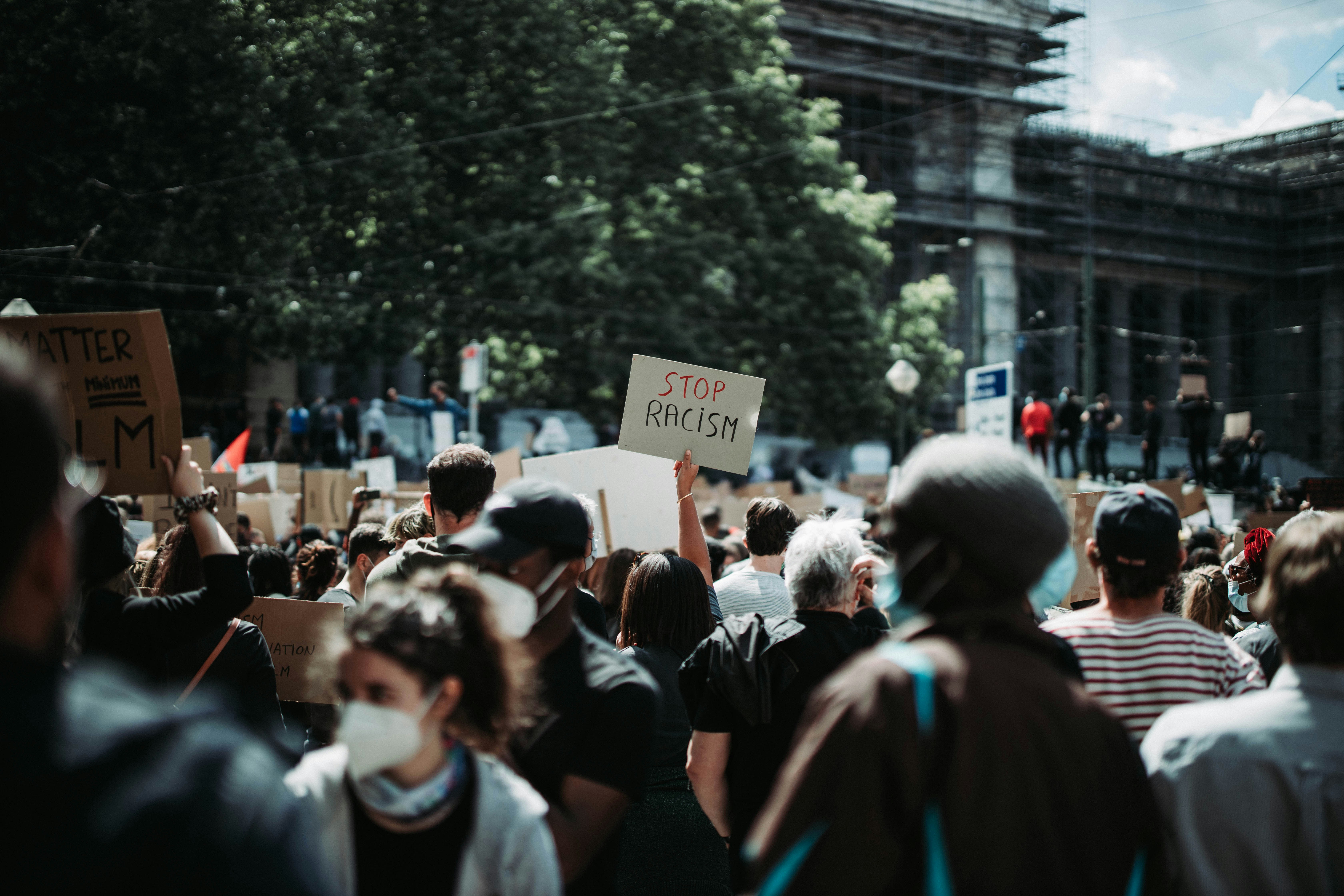 Protesters holding signs gather in a peaceful demonstration under dappled sunlight.