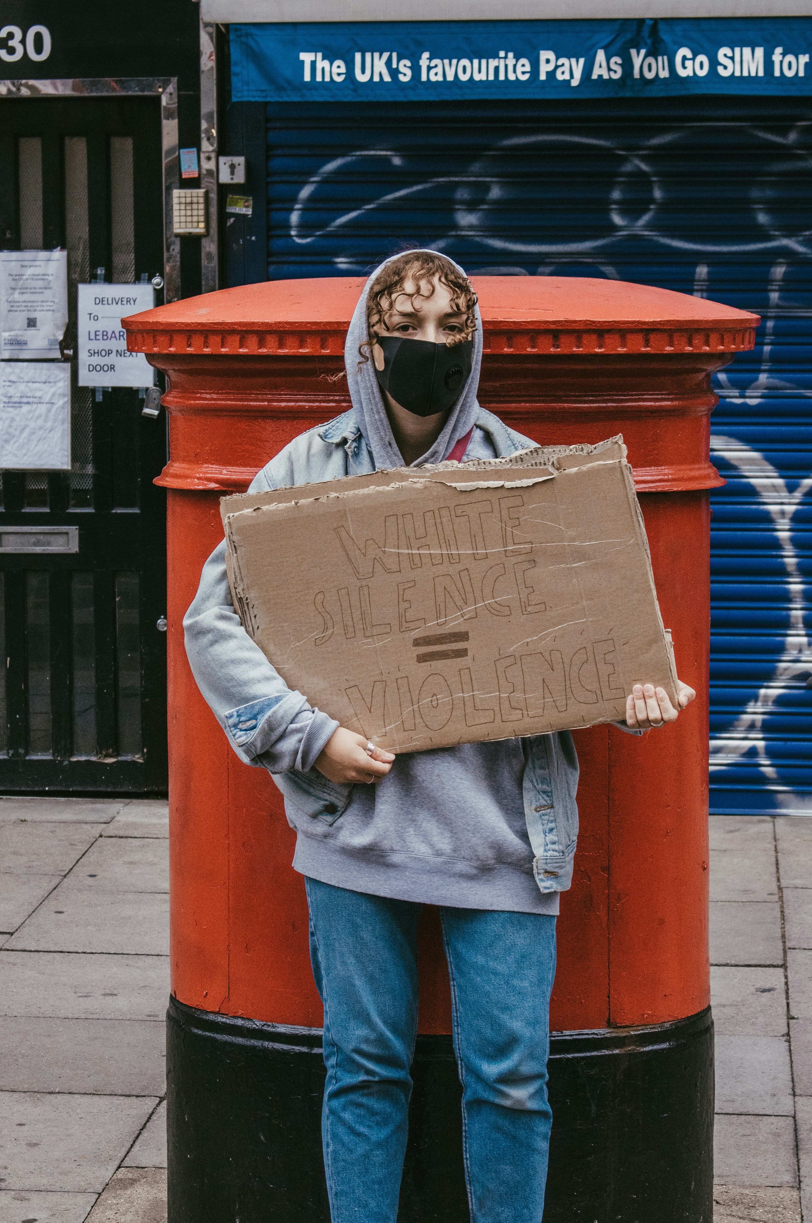 Protester holding a cardboard sign reading 'WHITE SILENCE = VIOLENCE' in front of a red postbox, emphasizing social justice themes.