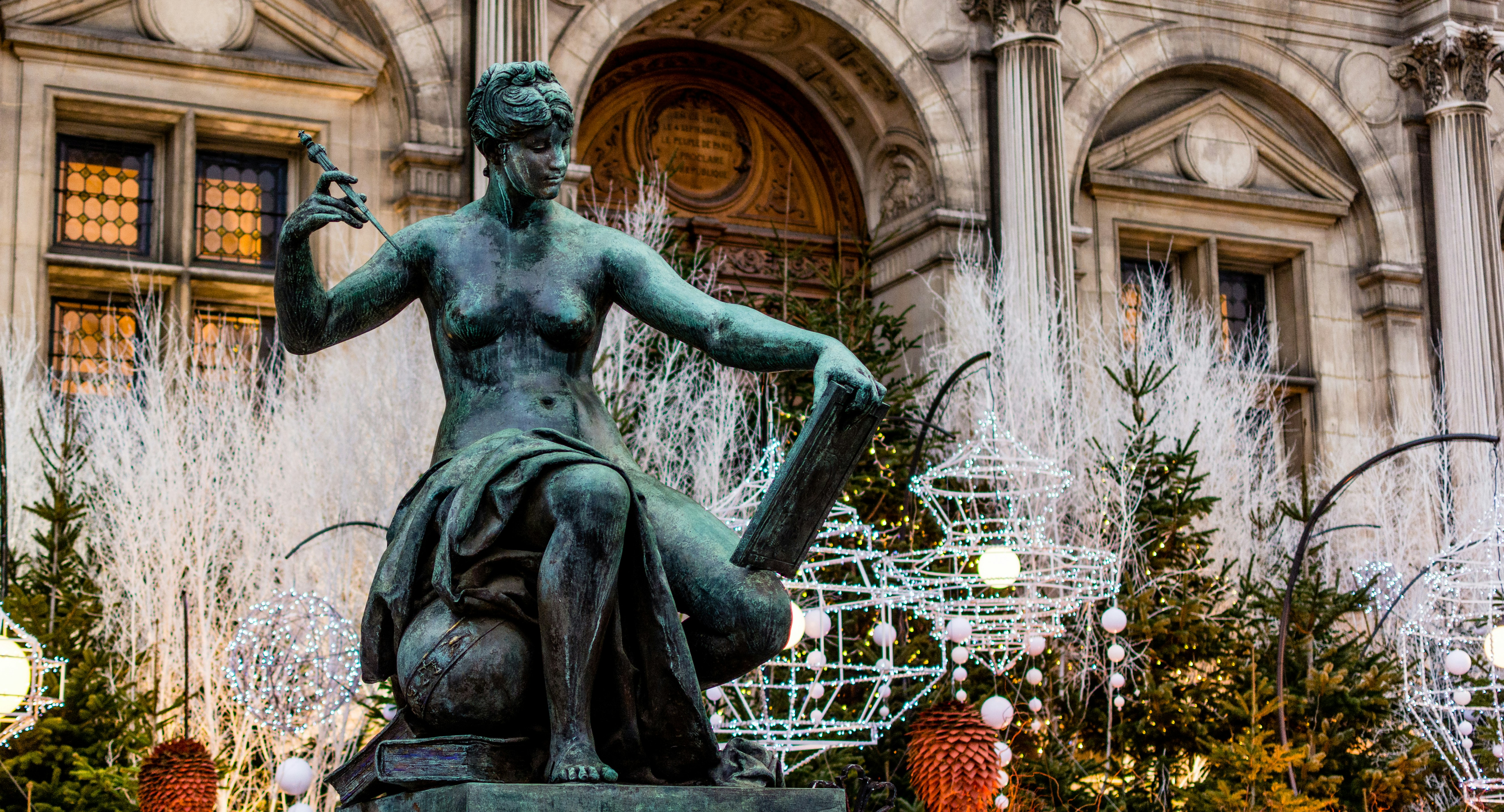 Bronze statue of a woman holding a book, surrounded by festive lights and greenery in a city square.
