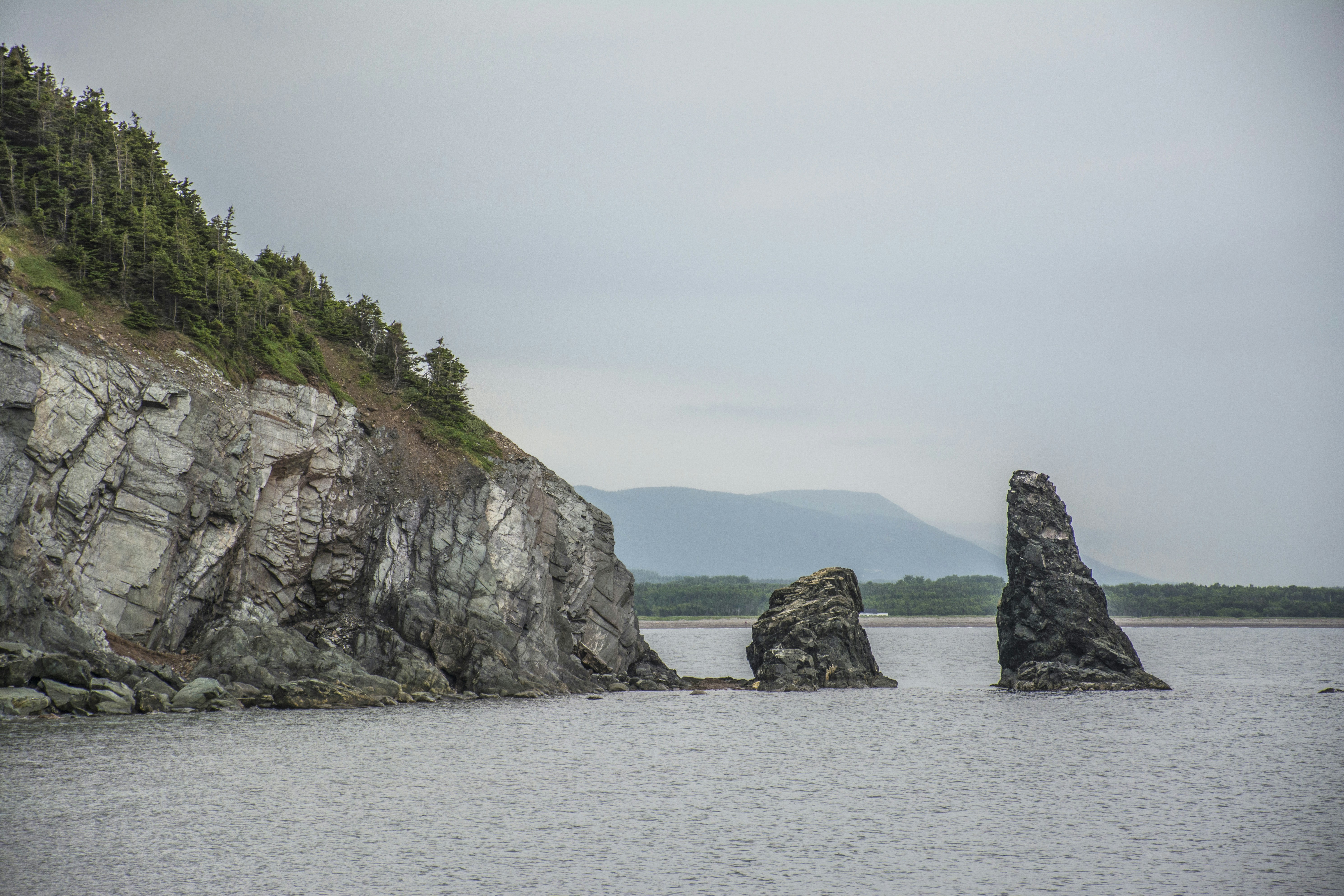 Rocky coastal cliffs with jagged formations extending into a calm sea under an overcast sky.
