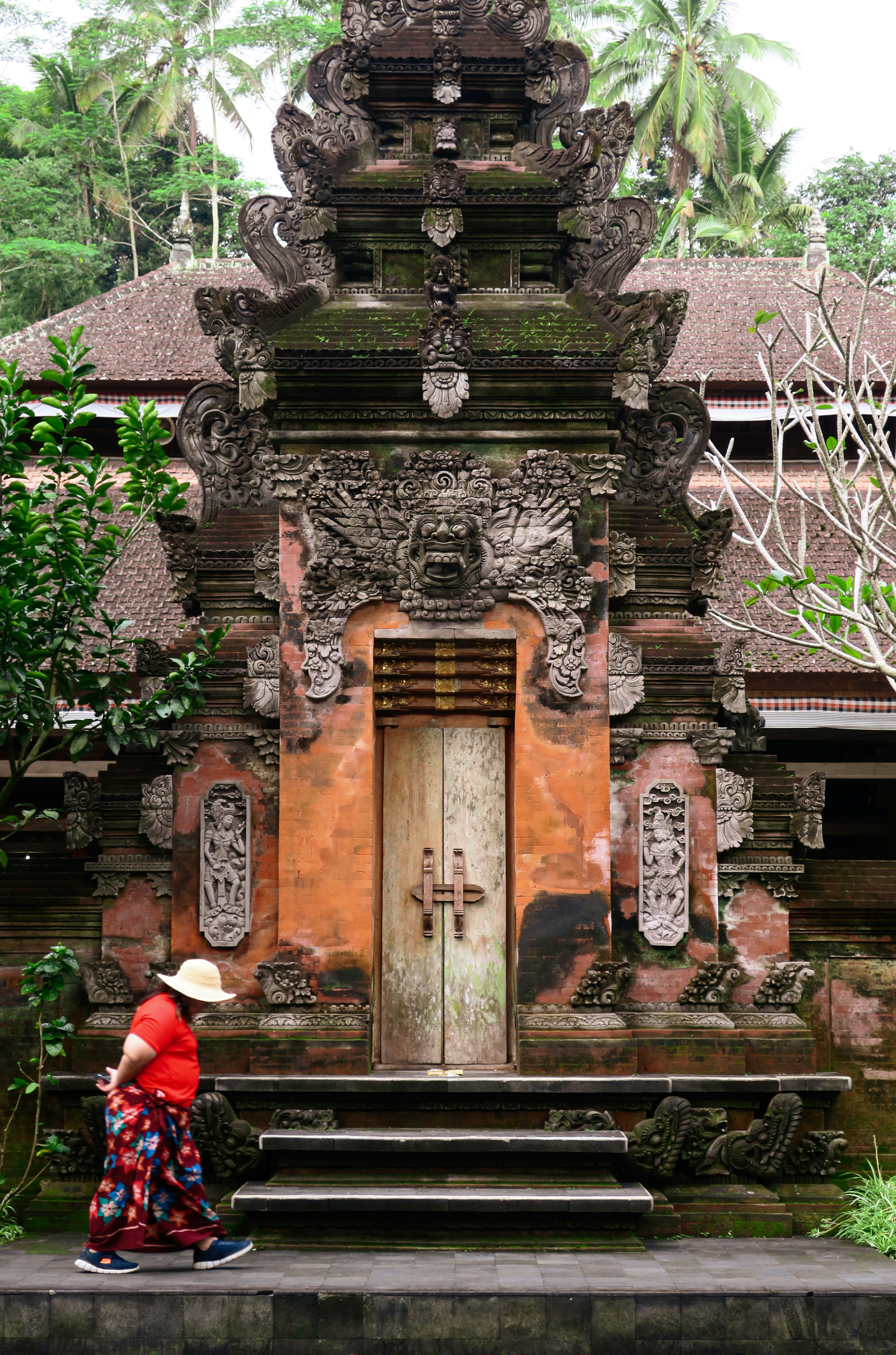 Intricately carved stone entrance with a figure in traditional attire walking by, surrounded by lush greenery. The scene reflects the rich cultural heritage of Bali.