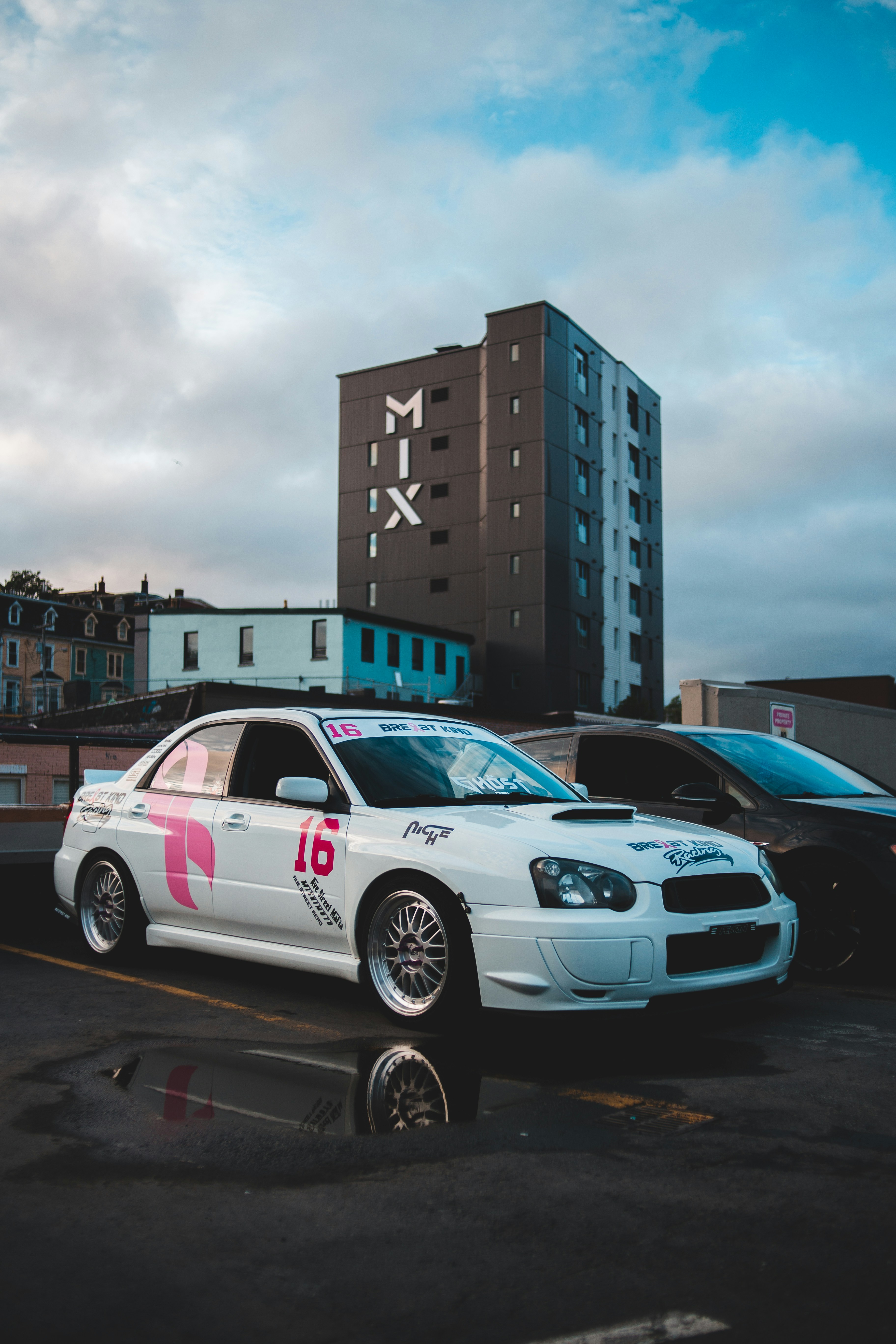 White race car with pink decals parked under a gray building at dusk
