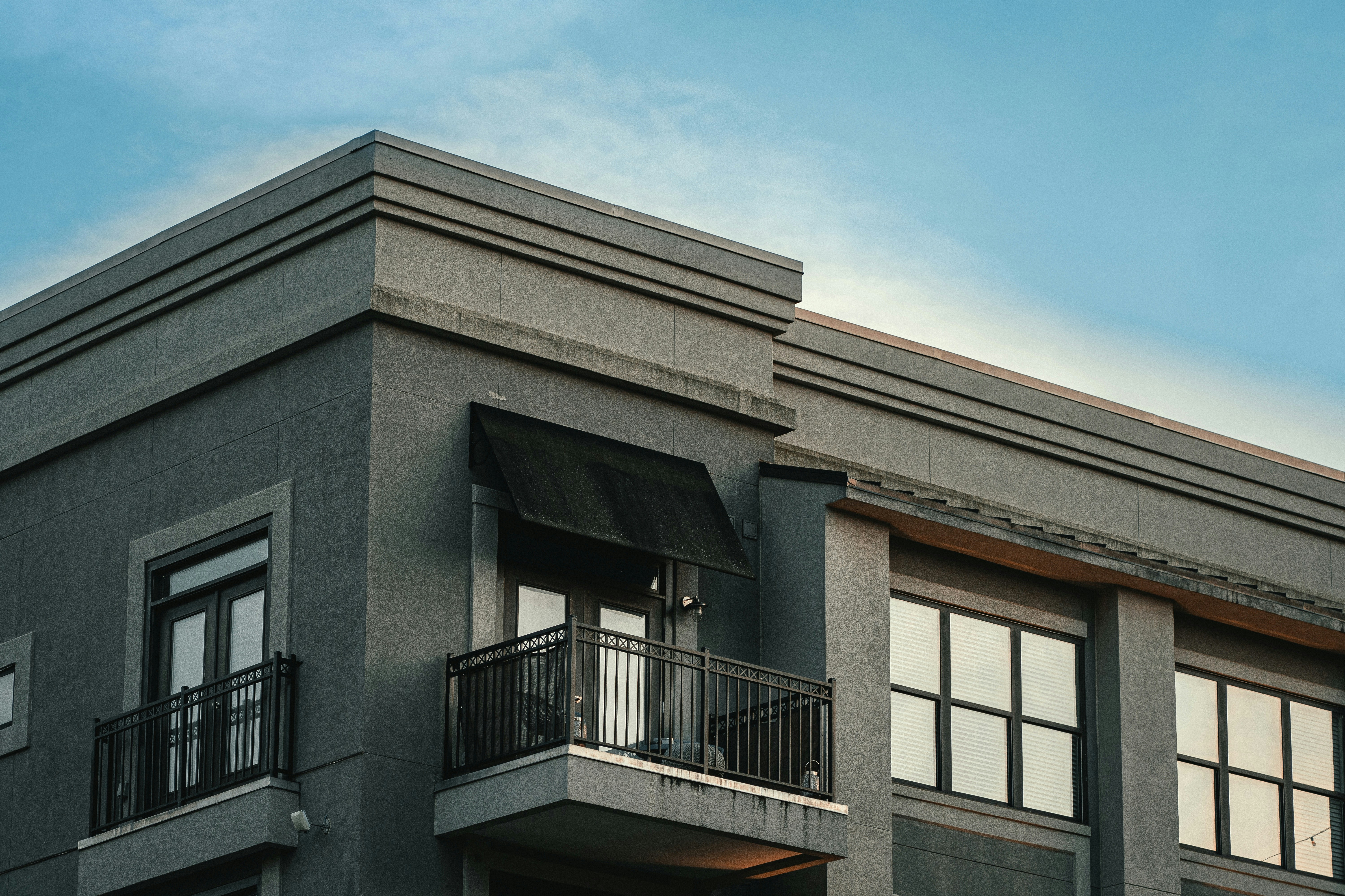 Architectural photograph of a grey building corner with a small balcony and black railing beneath a retractable awning. The clean lines and window pattern contrast with the blue sky.