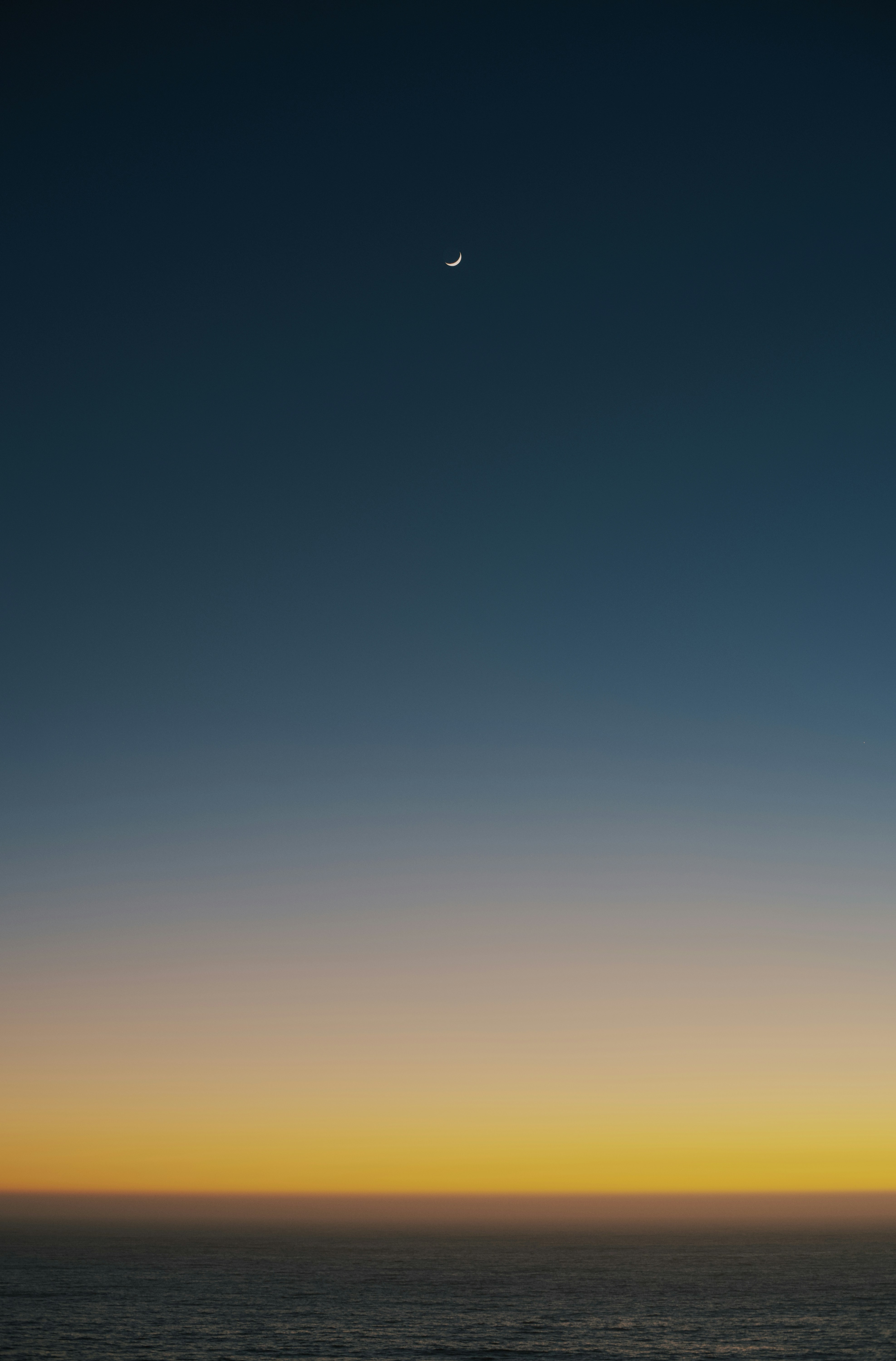 A crescent moon hangs delicately in a gradient sky transitioning from deep blue to warm orange near the horizon. The tranquil ocean reflects the serene colors of dusk.