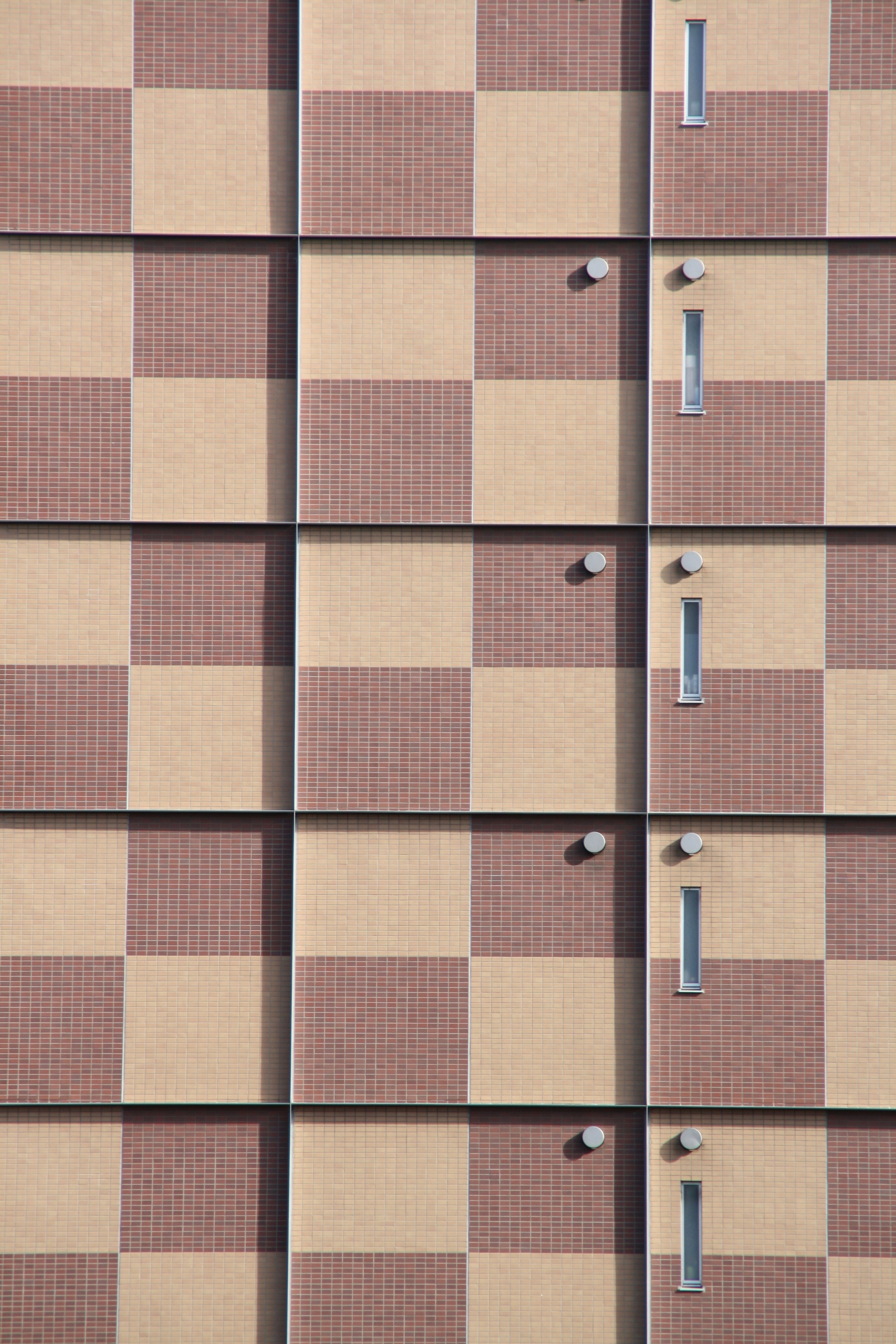 A close-up view of a building facade featuring an alternating pattern of beige and reddish-brown tiles, complemented by small windows and circular fixtures.