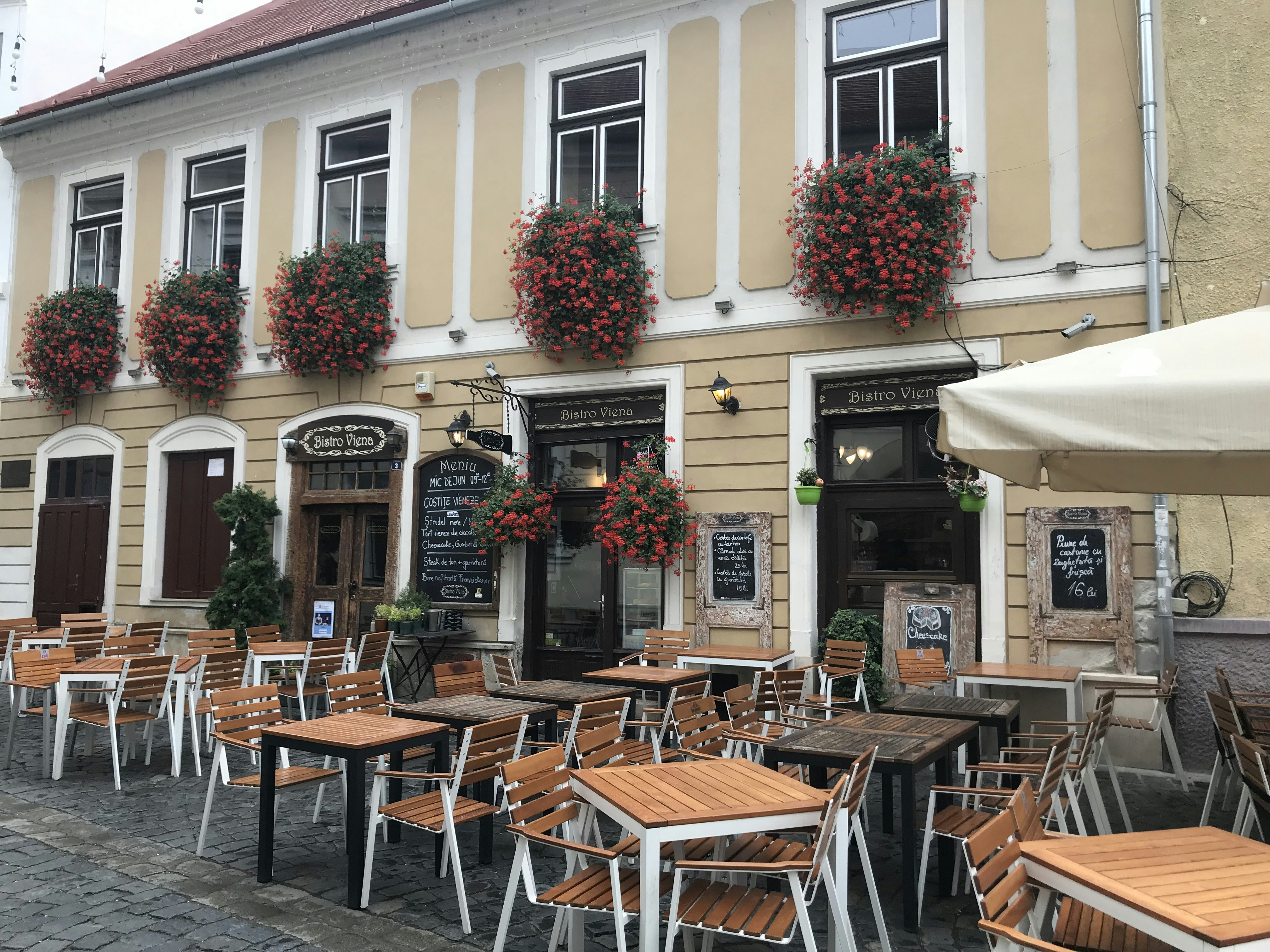 Quaint outdoor café with wooden tables and vibrant flower baskets adorning the façade of a historic building. Welcoming atmosphere invites patrons to enjoy a leisurely meal.
