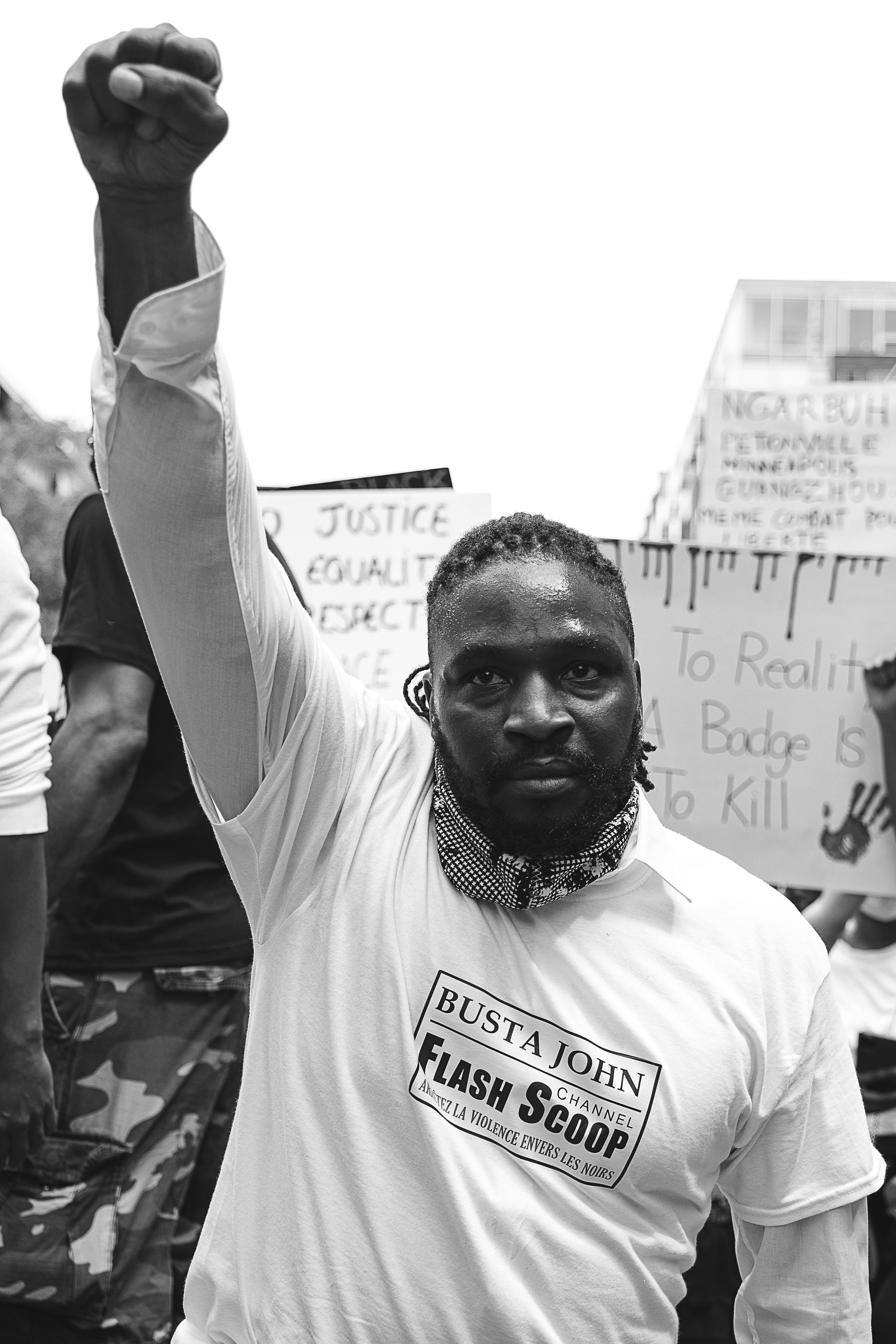 Man in a white t-shirt raising his fist at an anti-racism protest, surrounded by signs advocating for justice and equality.