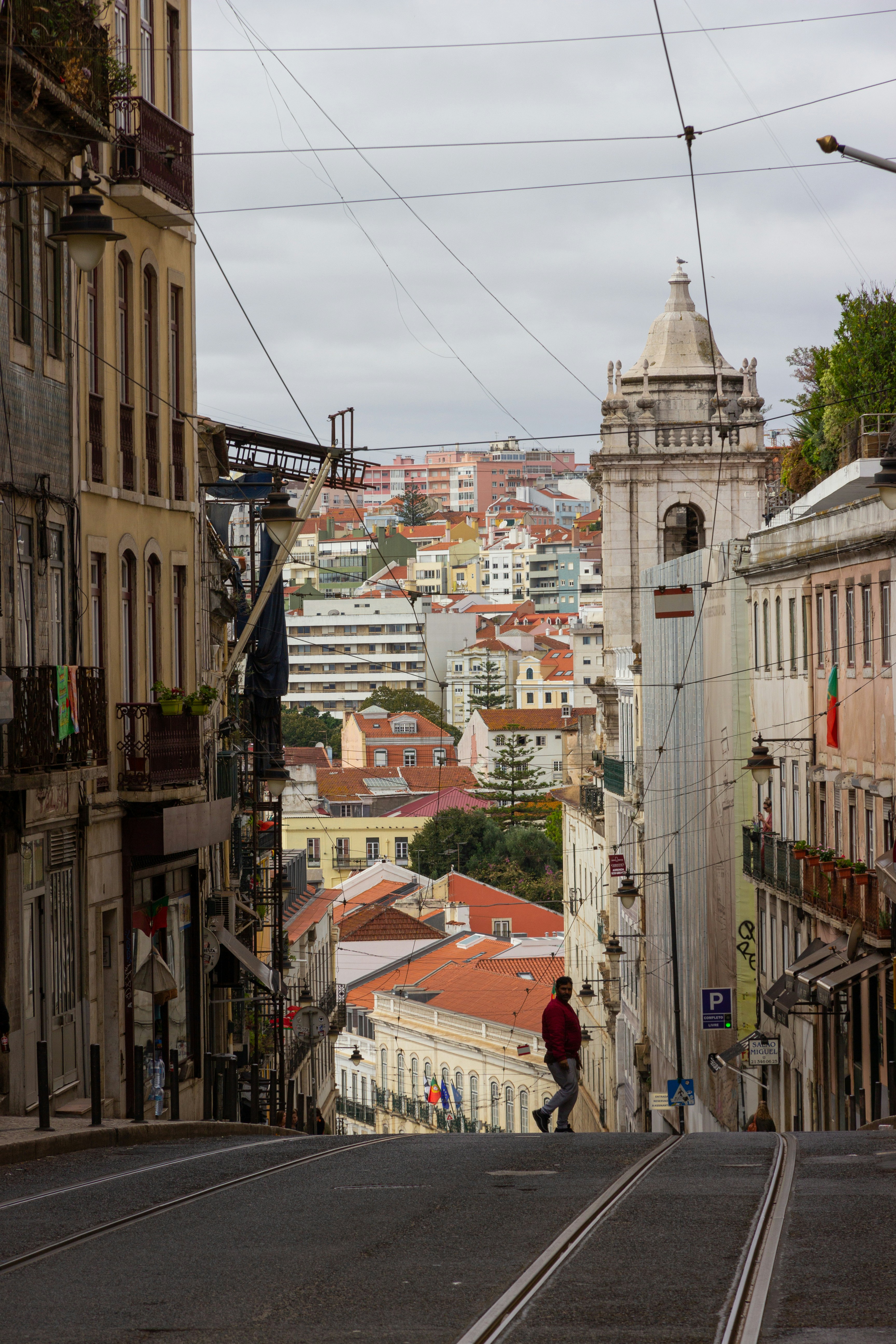 A pedestrian traverses a sloped street lined with historic buildings, capturing the essence of Lisbon's vibrant city life. The distant architecture adds depth to the urban landscape.