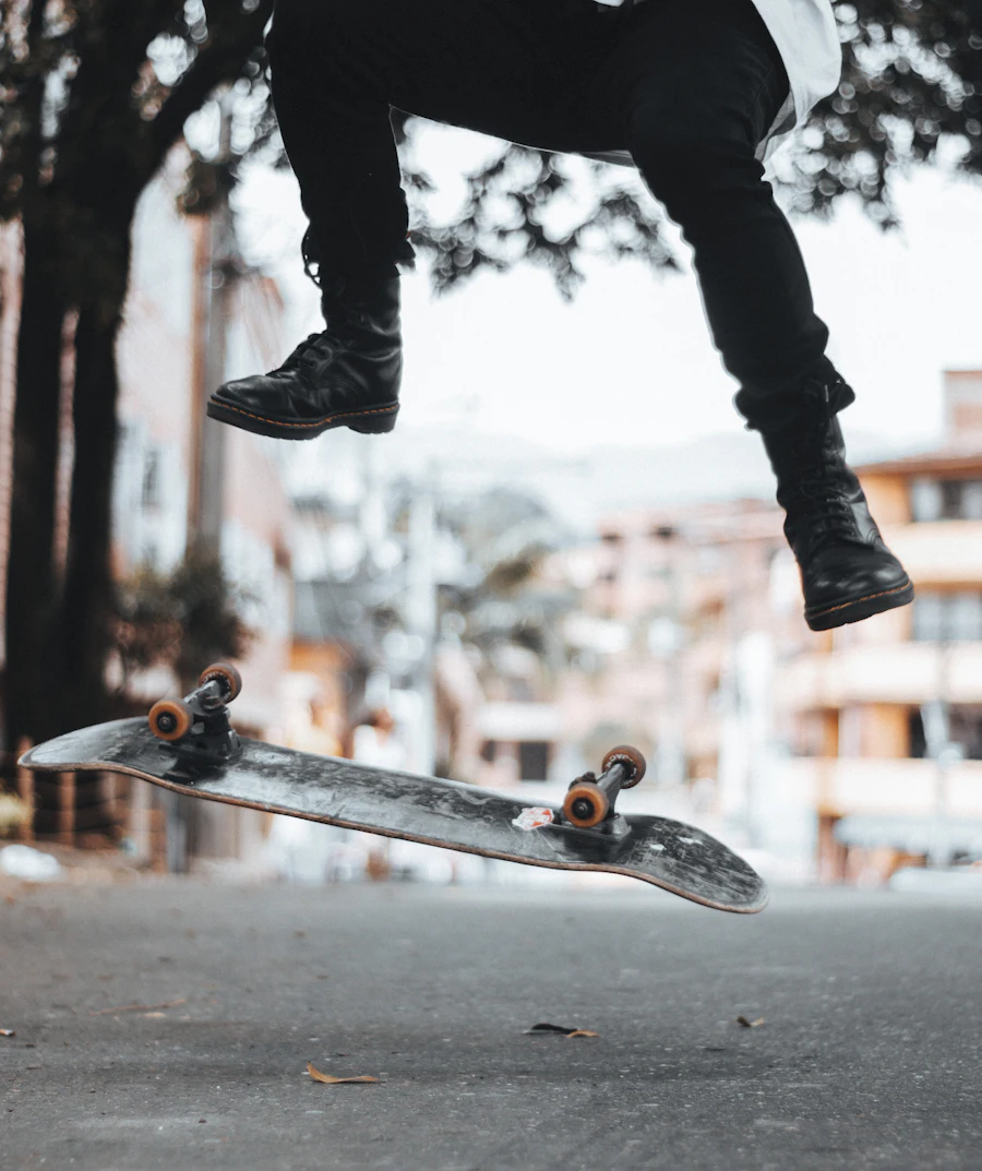 A skateboard spins above the pavement while a rider’s boots hang in the air overhead, with a softly blurred city street and trees in the background.