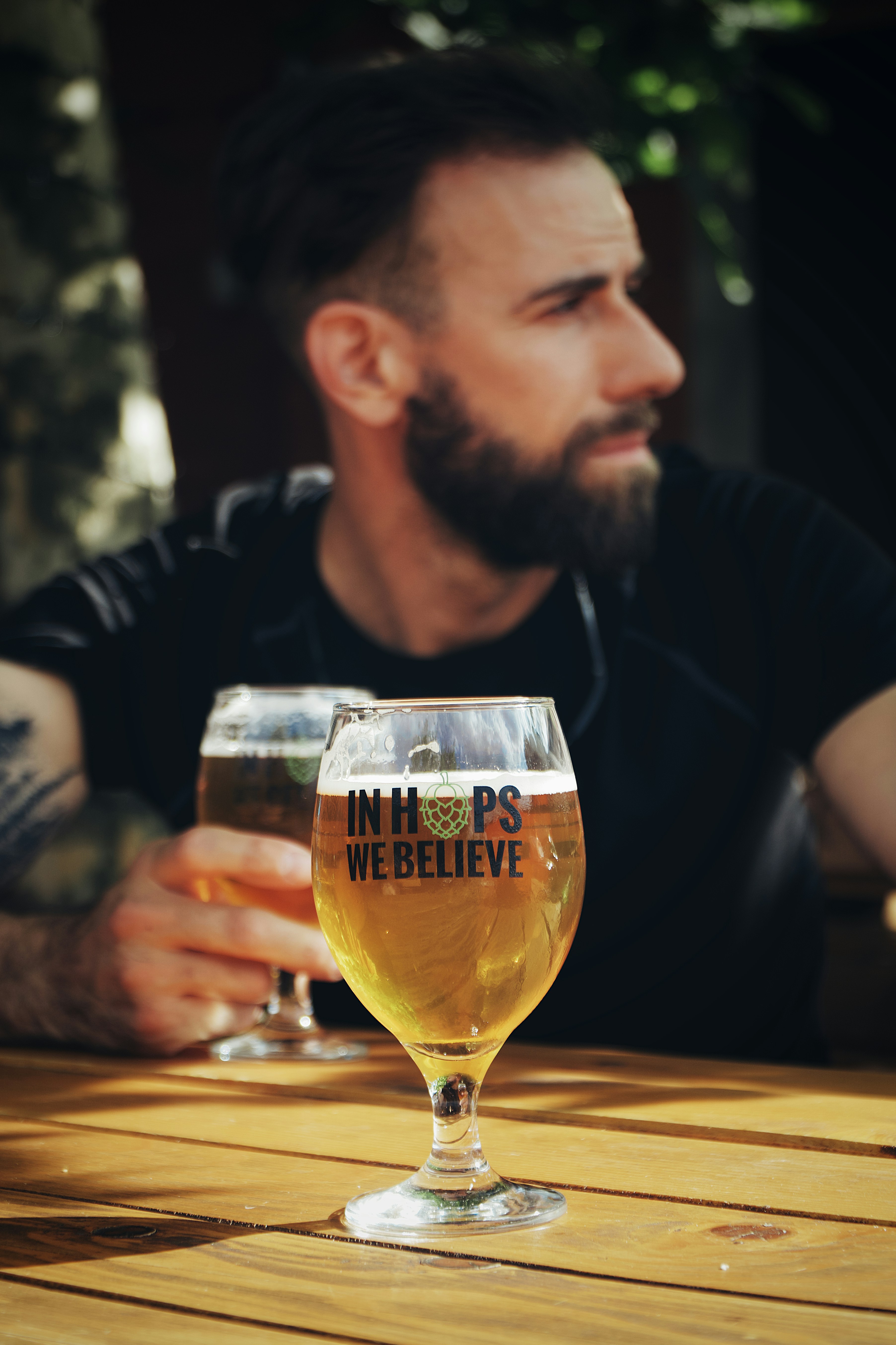 Man holding a beer glass emblazoned with 'In Hops We Believe' in a sunlit setting.