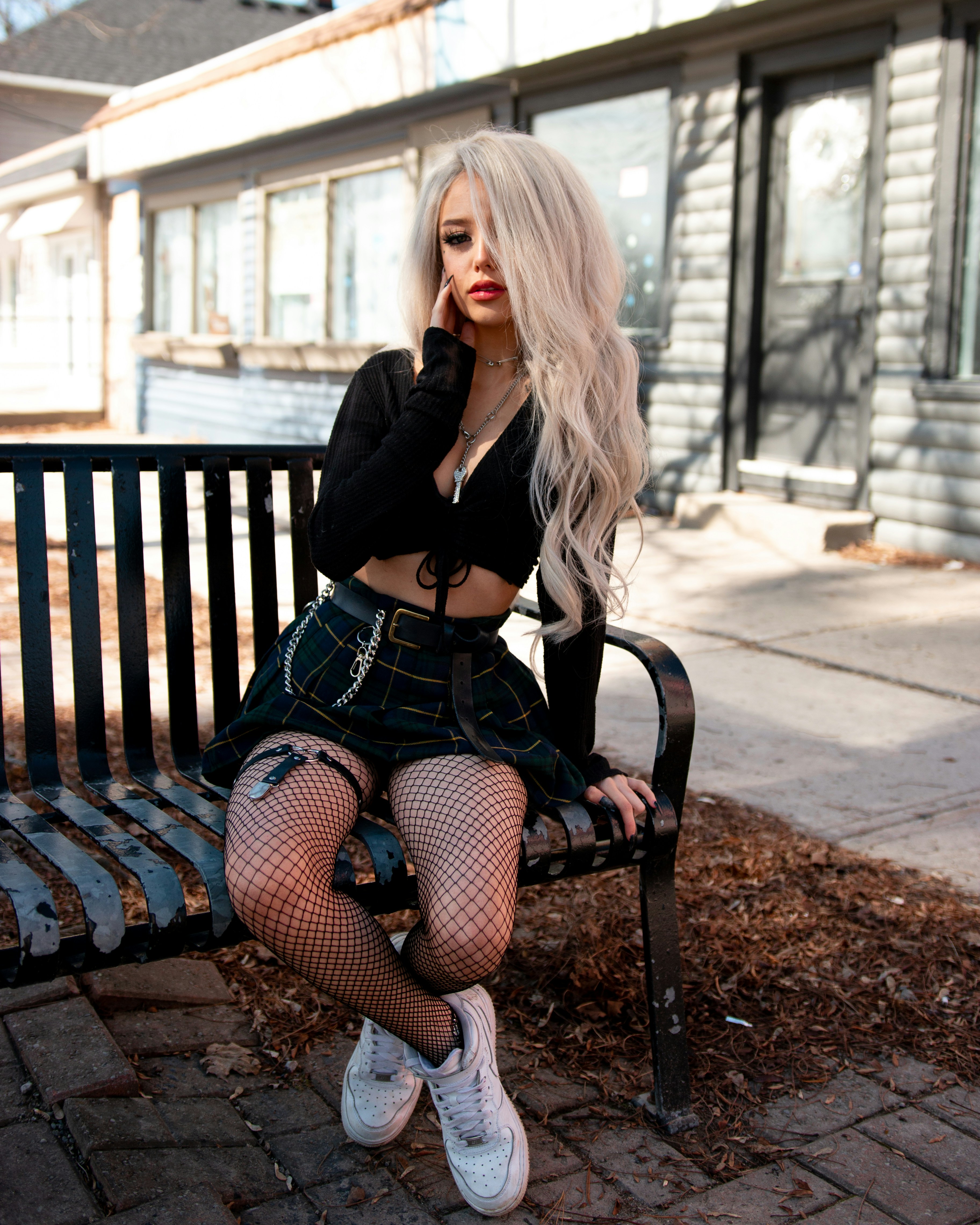 A stylish individual in a black crop top and plaid skirt sits on a park bench, showcasing a blend of urban fashion and casual comfort.