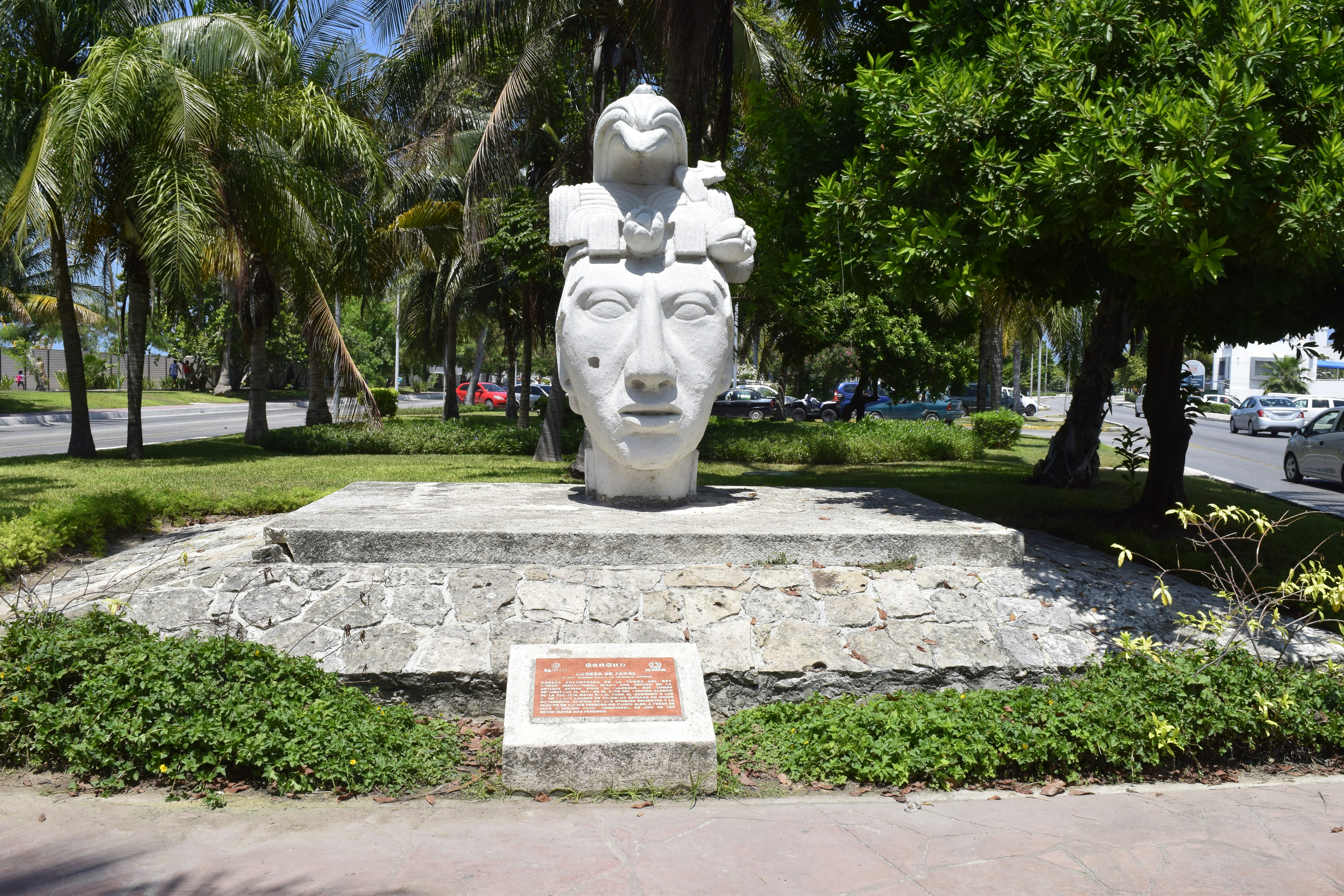 A striking white sculpture featuring a human head with a bird atop, surrounded by lush greenery and a stone base, conveying cultural significance.