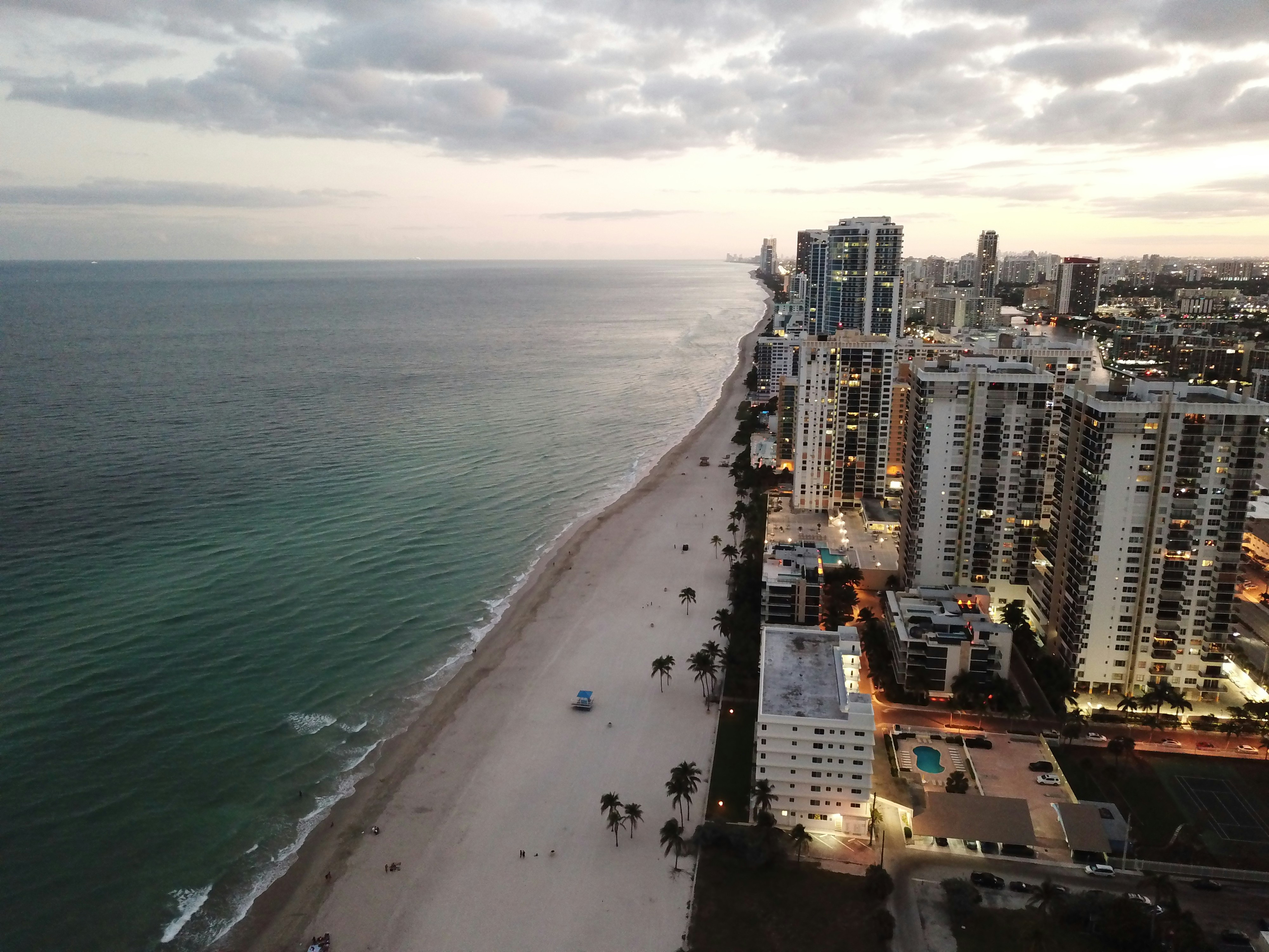 Aerial view of a coastal city with high-rise buildings lining a sandy beach as the sun sets.