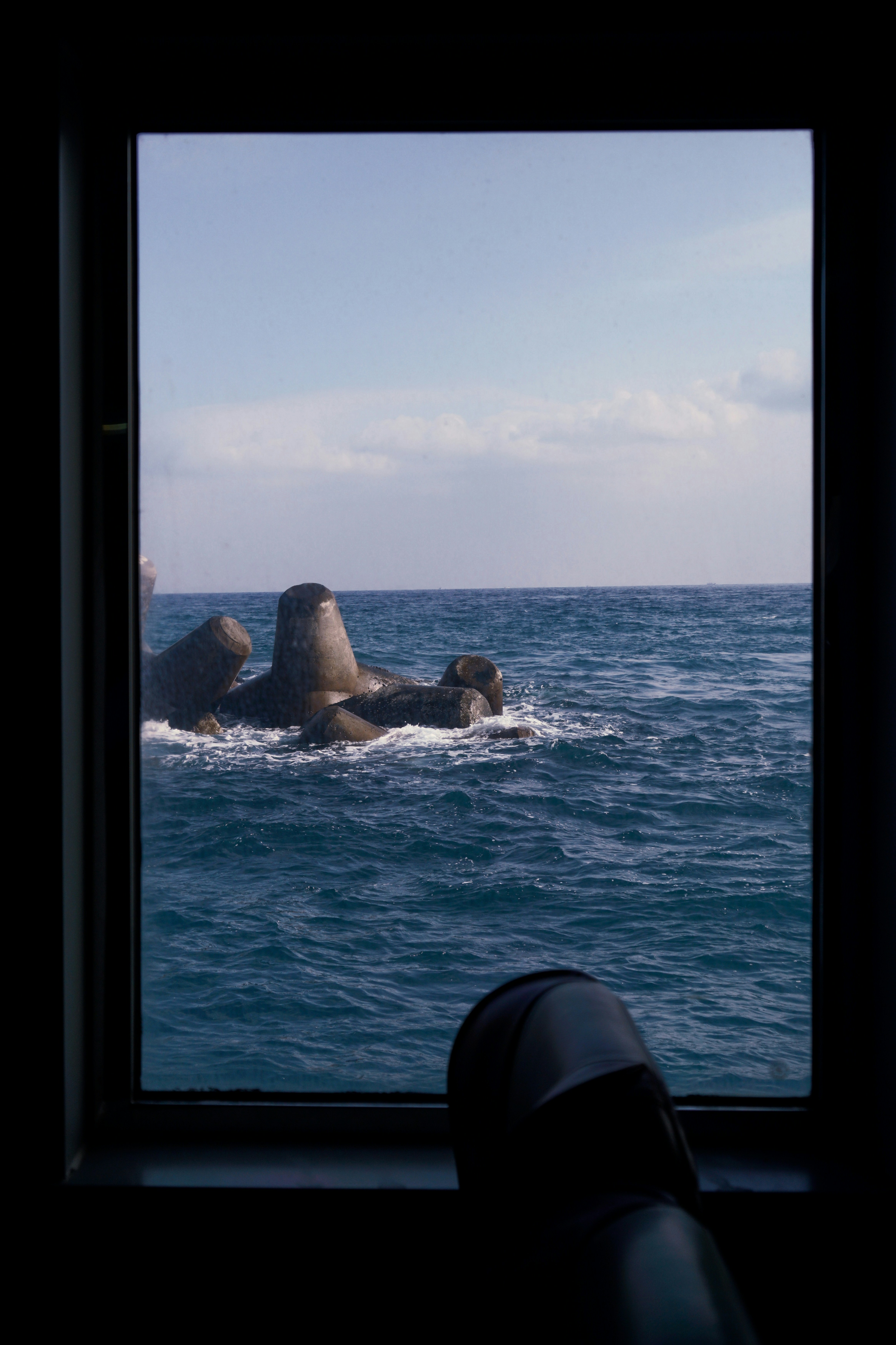View of rocky formations rising from the ocean, captured through a window frame. Gentle waves lap against the stones.