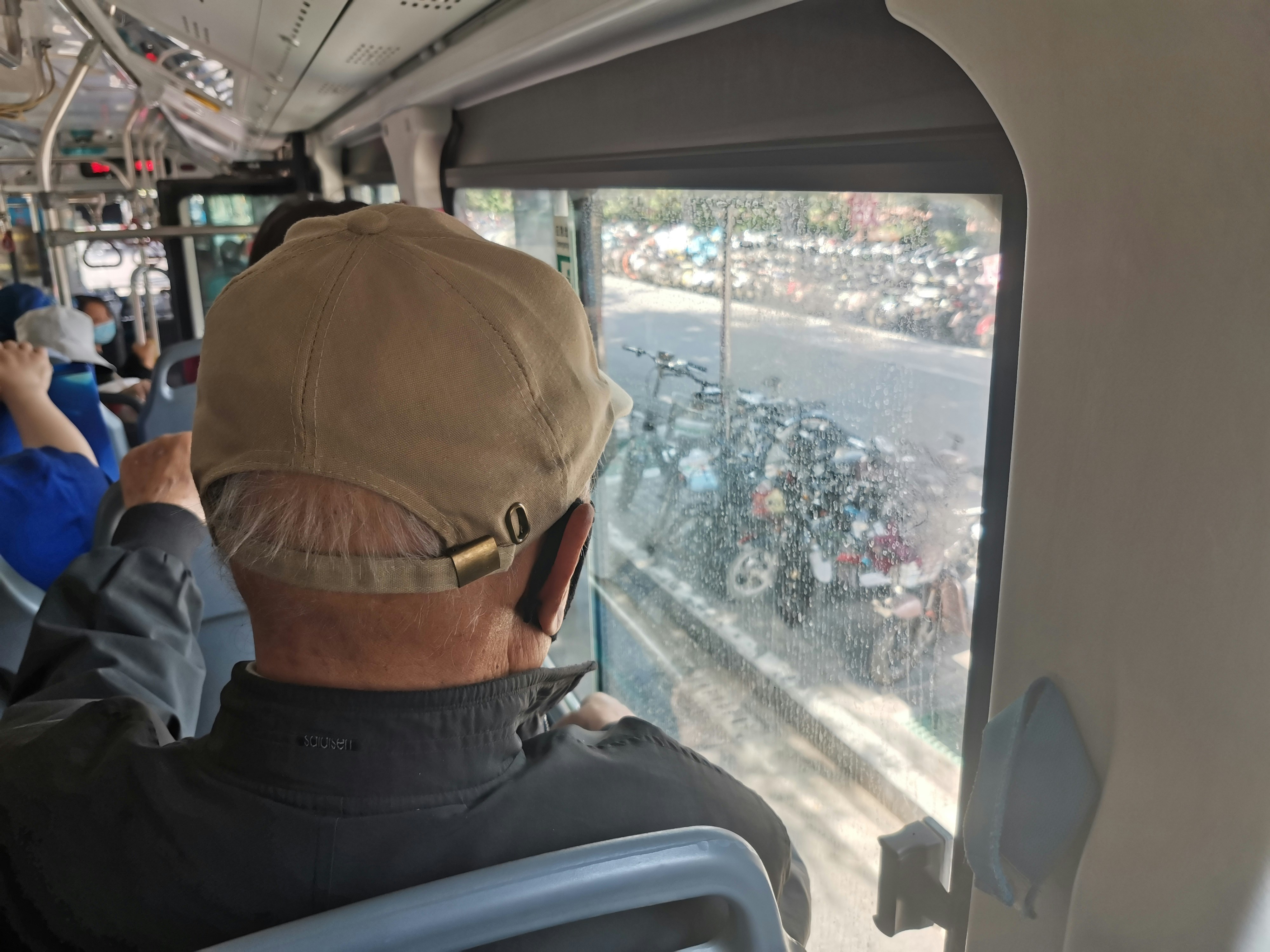 Back of a commuter caped head looking out a rain-splattered bus window
