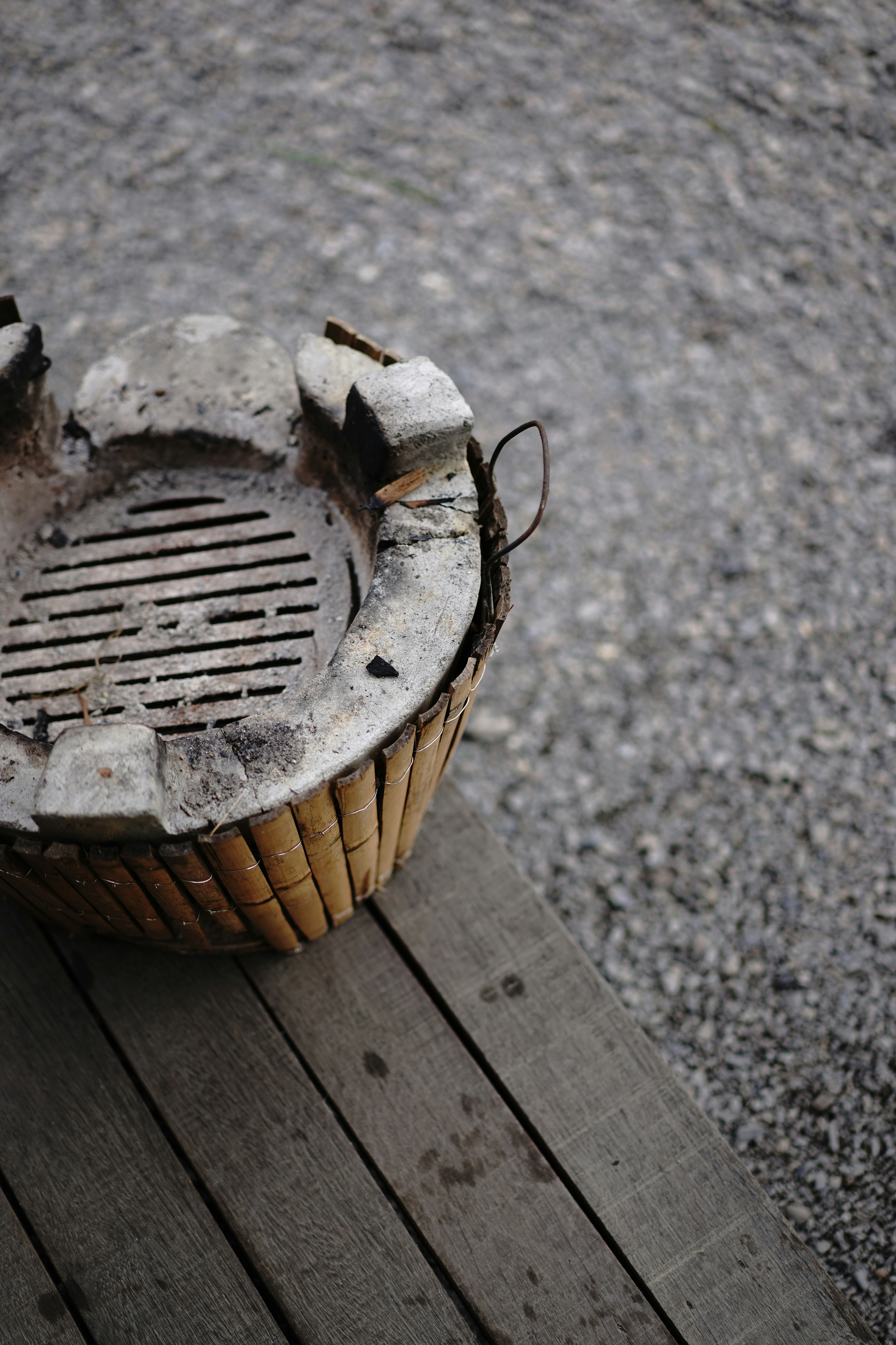Weathered charcoal grill made from bamboo and stone, showcasing signs of use and age. The grill sits on a wooden surface amidst a gravel backdrop.