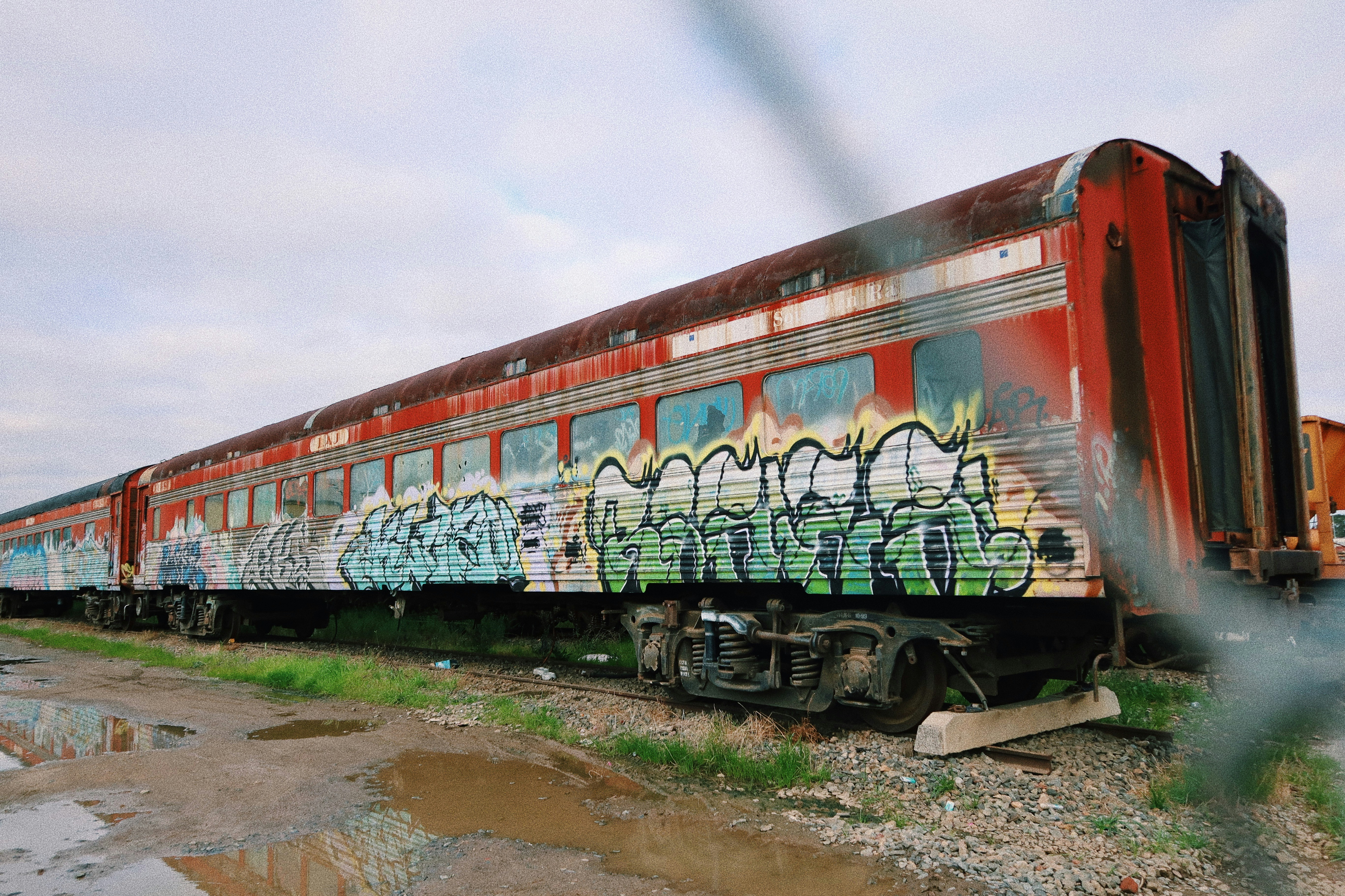 Graffiti-covered red railcar stretches along weathered tracks in a muddy rail yard under a cloudy sky. Bold colors and lettering sweep the car’s side while the blurred foreground hints at the camera’s close approach.