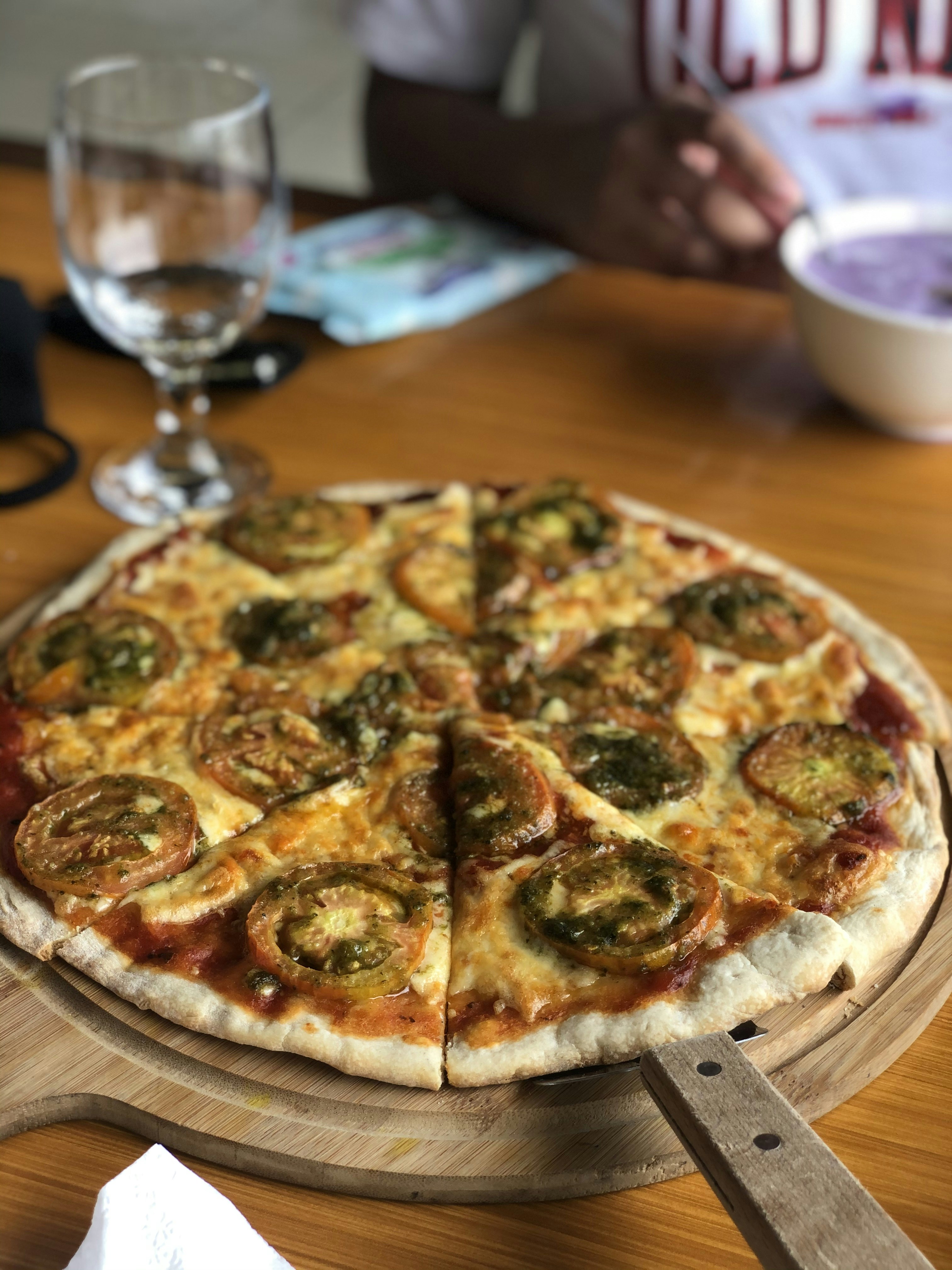 Delicious pizza topped with fresh green peppers and golden cheese, served on a wooden platter. A glass of water and a bowl of dessert are visible in the background.