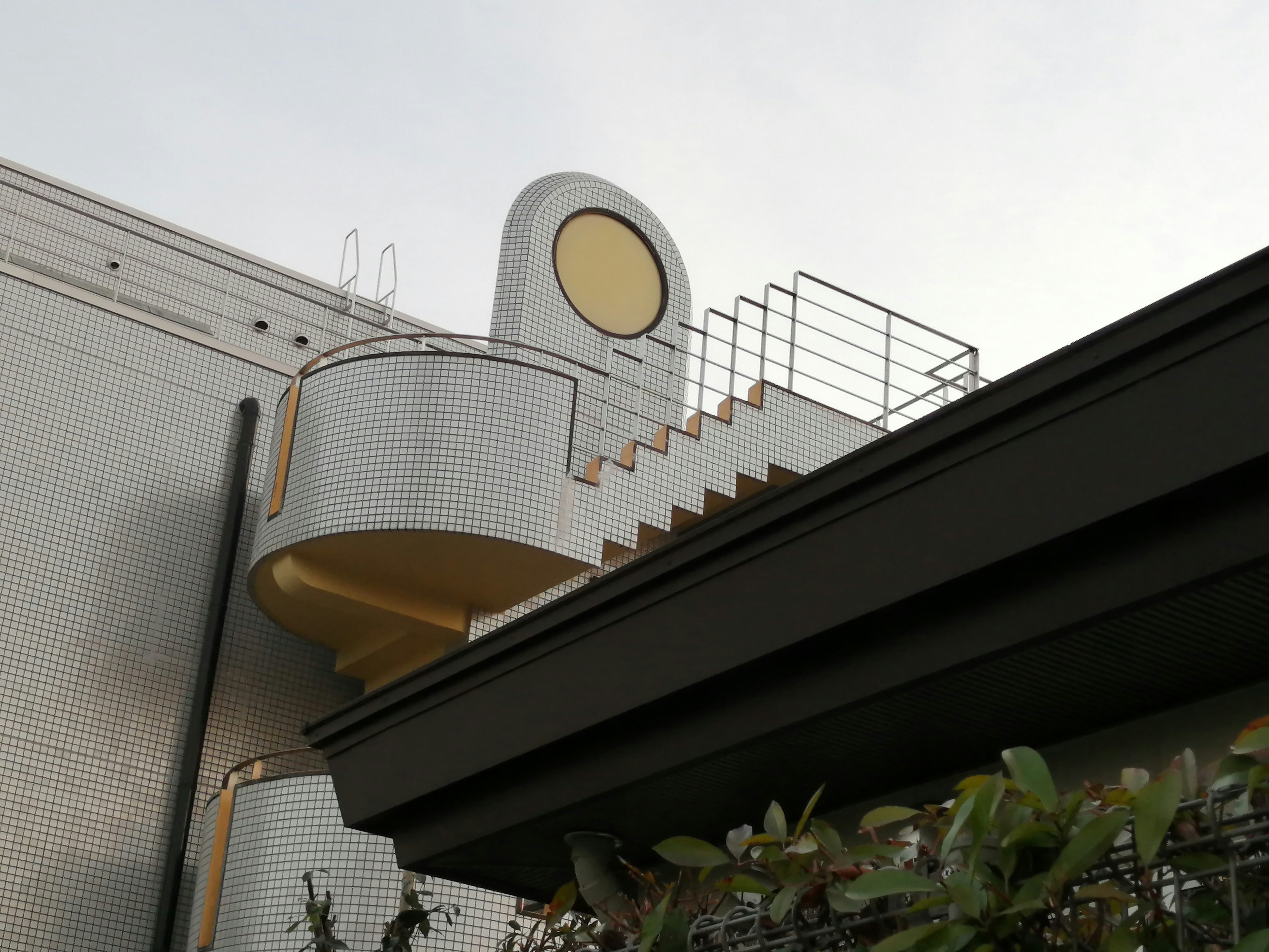 Photo of a tiled building façade with a prominent yellow circular feature, a curved balcony, and a dark foreground overhang.