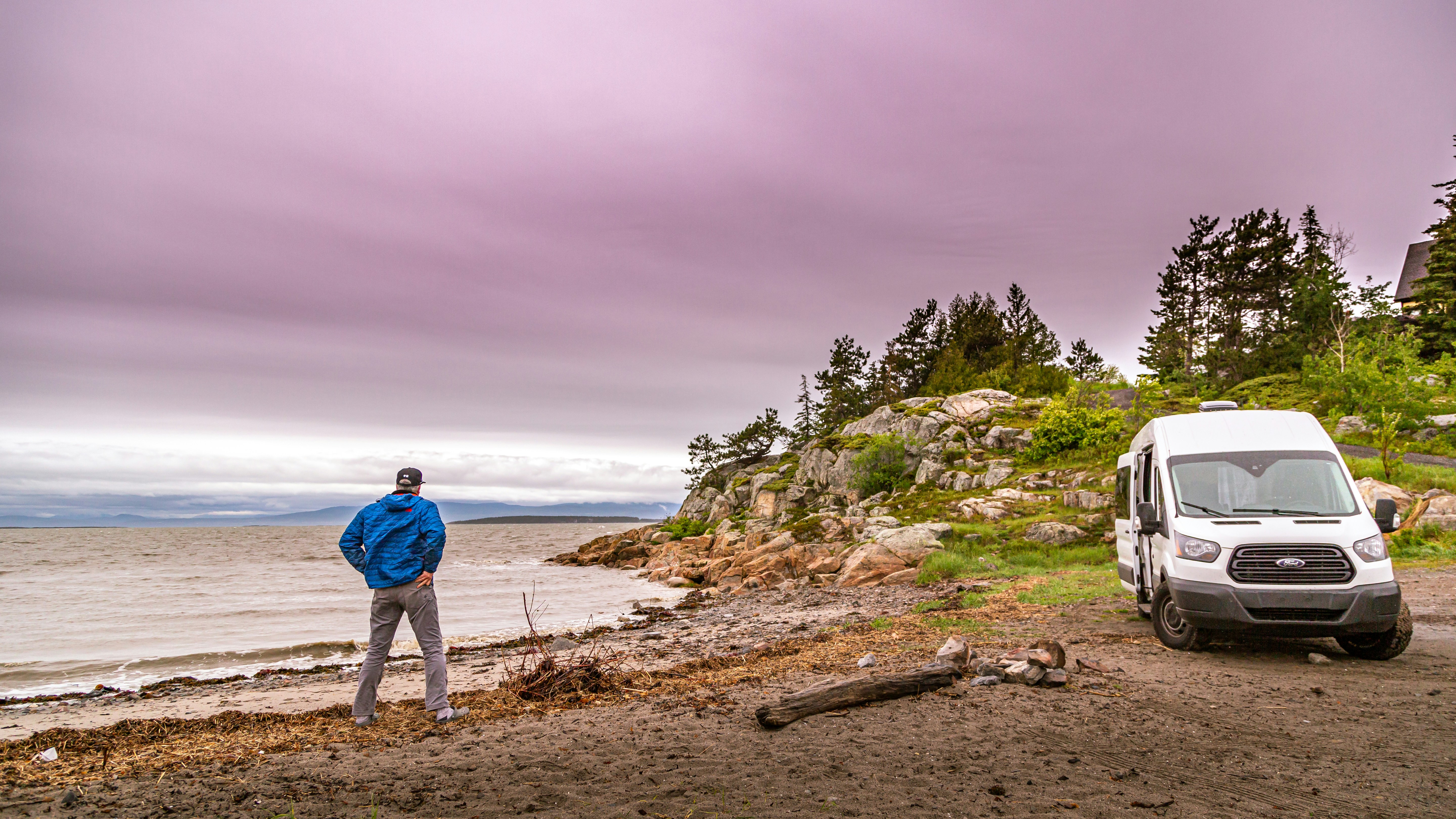 A person in a blue jacket gazes out over a calm sea, with a camper van parked nearby and rocky cliffs in the background.