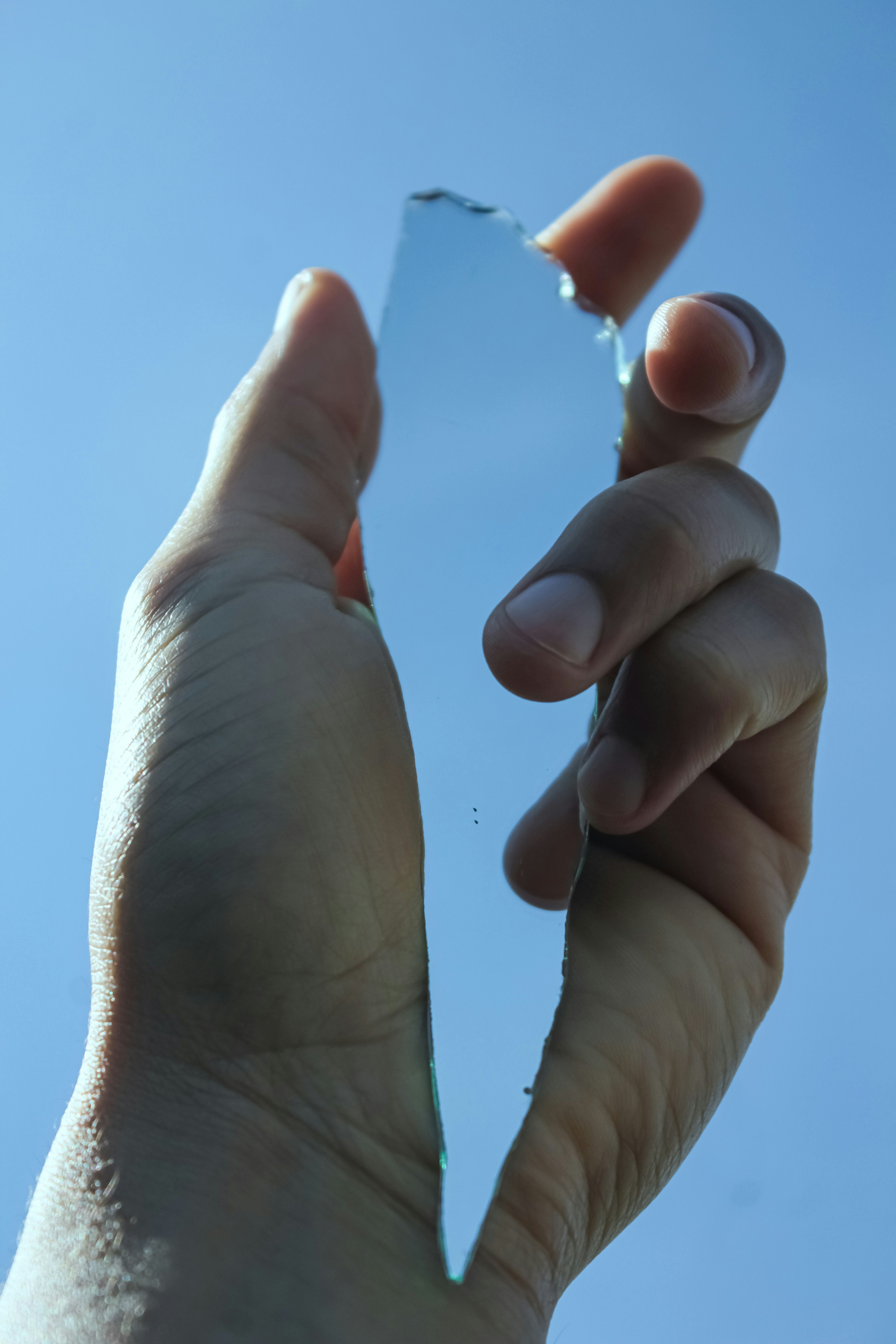 A hand holds a shard of glass against a clear blue sky, capturing reflections and light. The contrast emphasizes the fragility of the glass and the vastness of the sky.