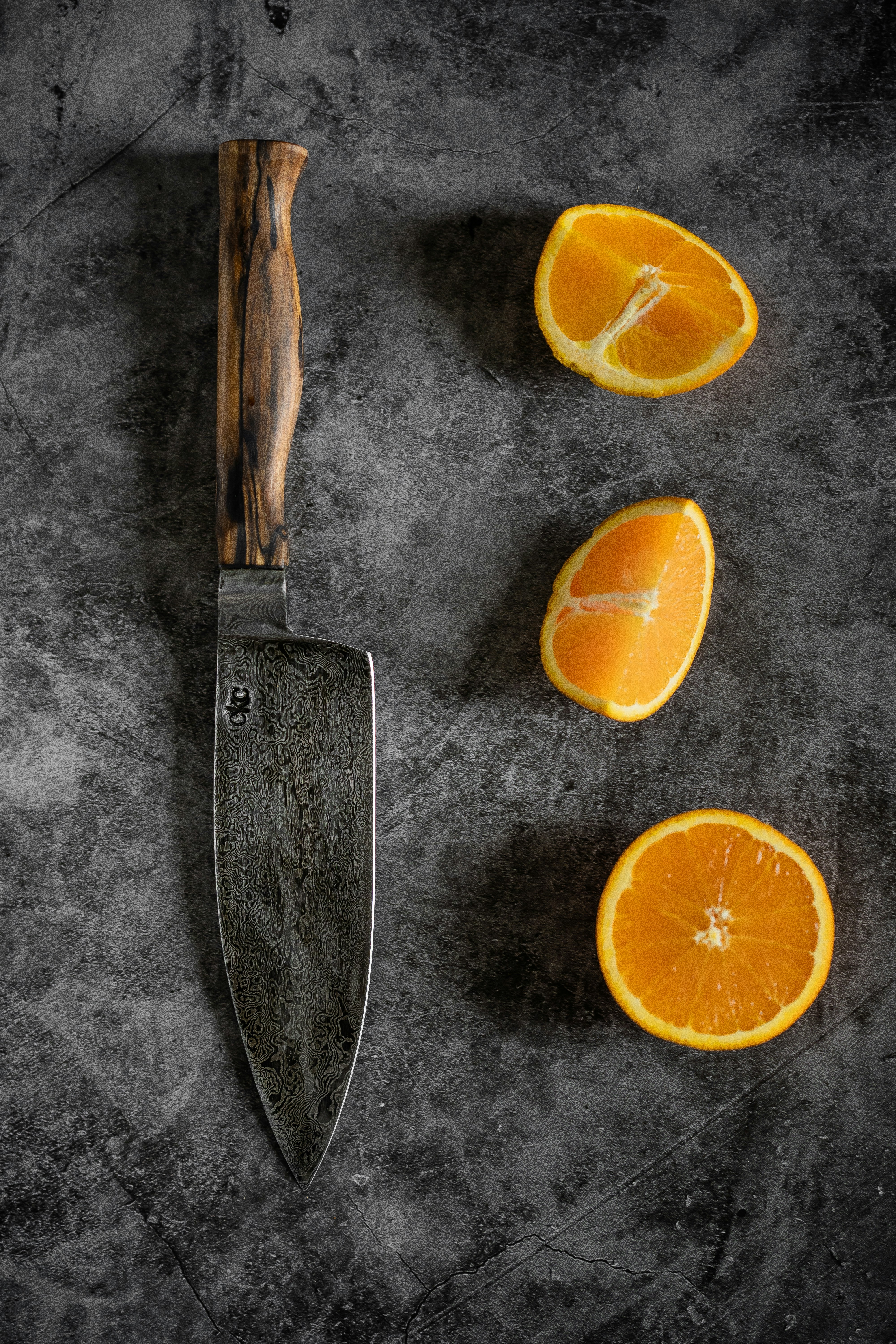 A finely crafted kitchen knife rests beside sliced oranges on a textured gray surface, showcasing a blend of culinary artistry and fresh ingredients.
