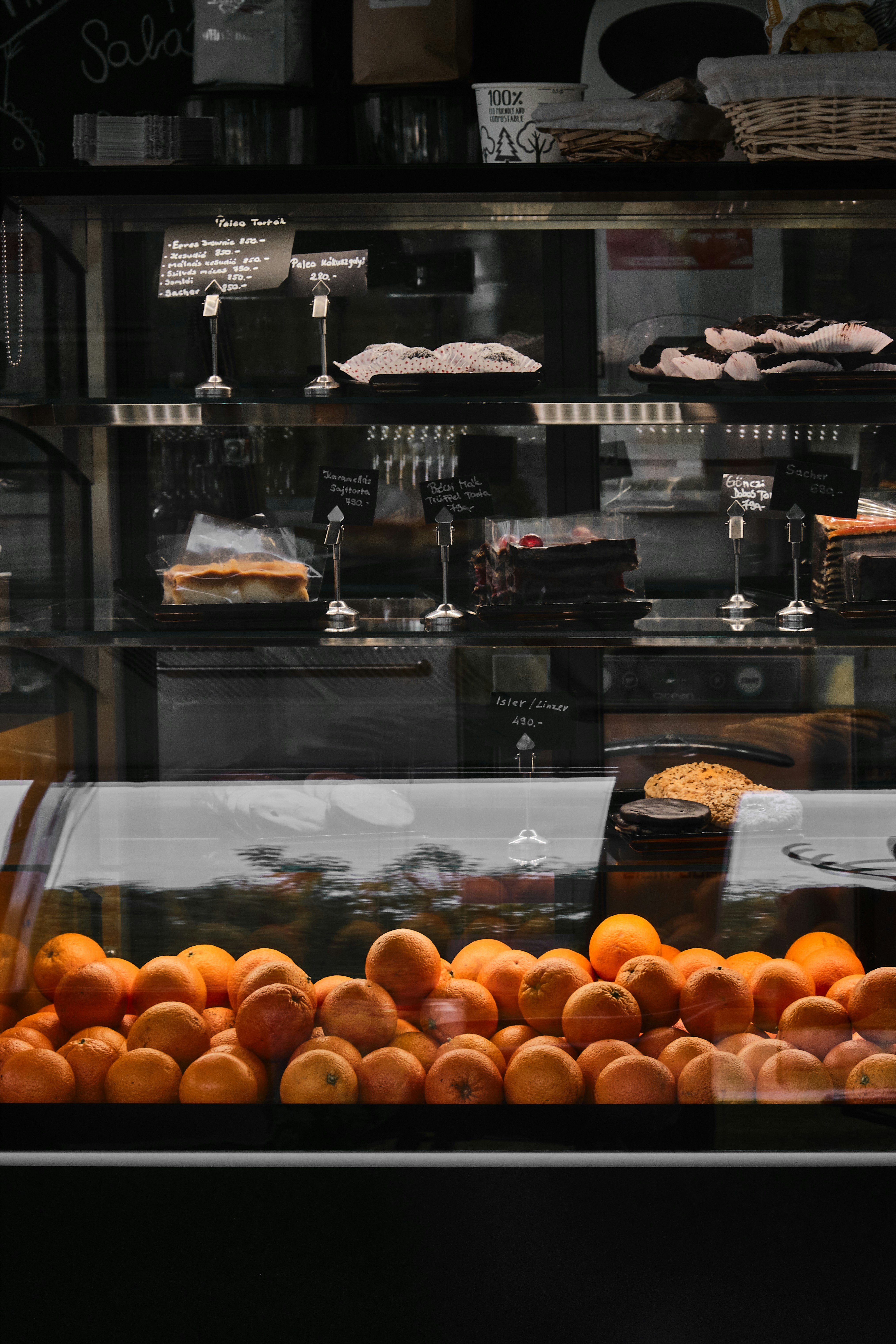 Vibrant oranges displayed prominently in a glass case, surrounded by an array of pastries and desserts. The scene captures the essence of a cozy café ambiance.