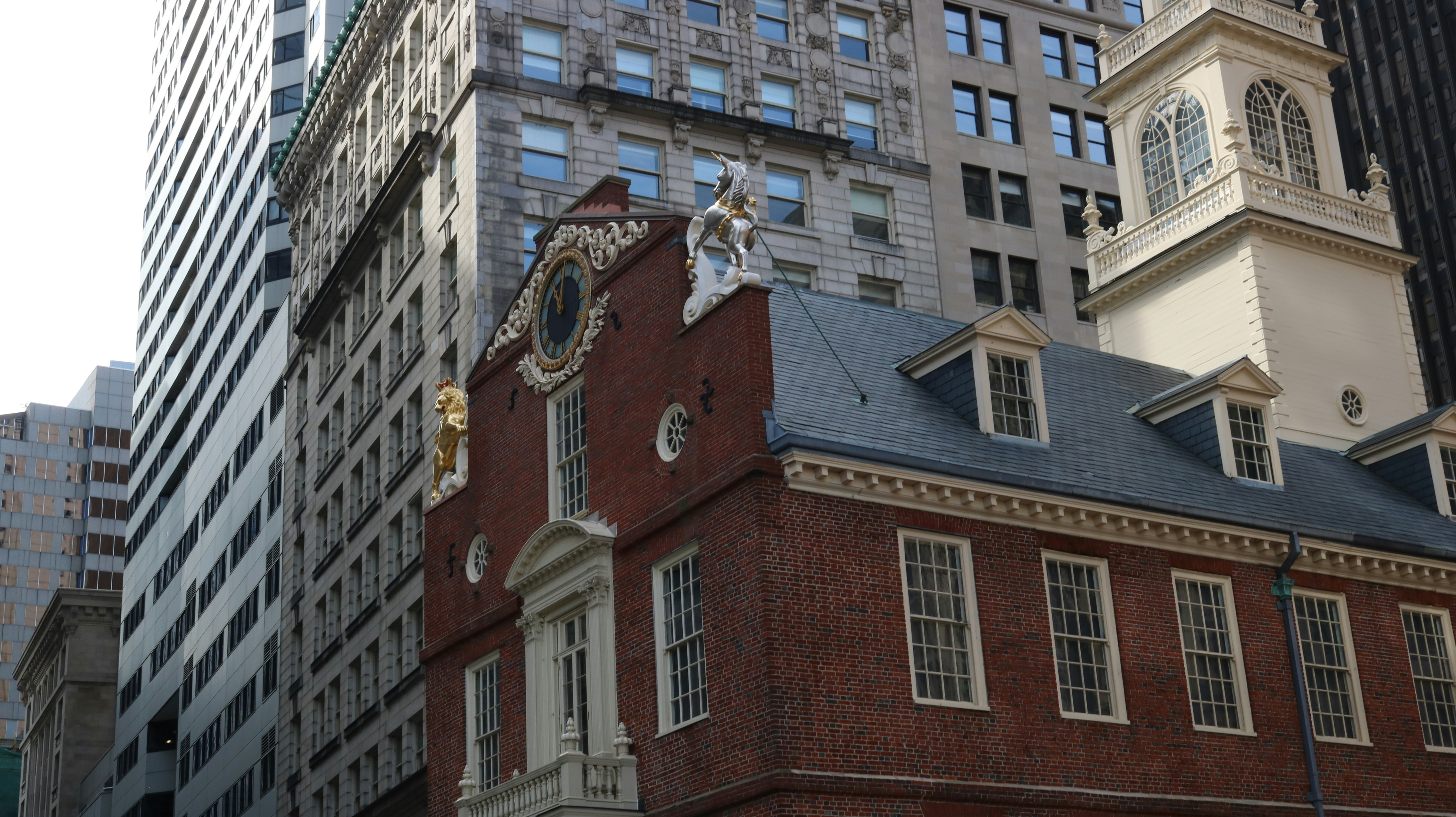 Historic brick building with ornate details nestled between towering skyscrapers in an urban setting.