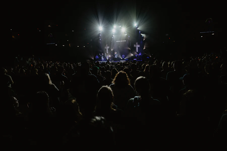 Dark concert venue packed with audience silhouettes facing a brightly lit stage. Starburst spotlights and cross-shaped stage elements glow above the performers.