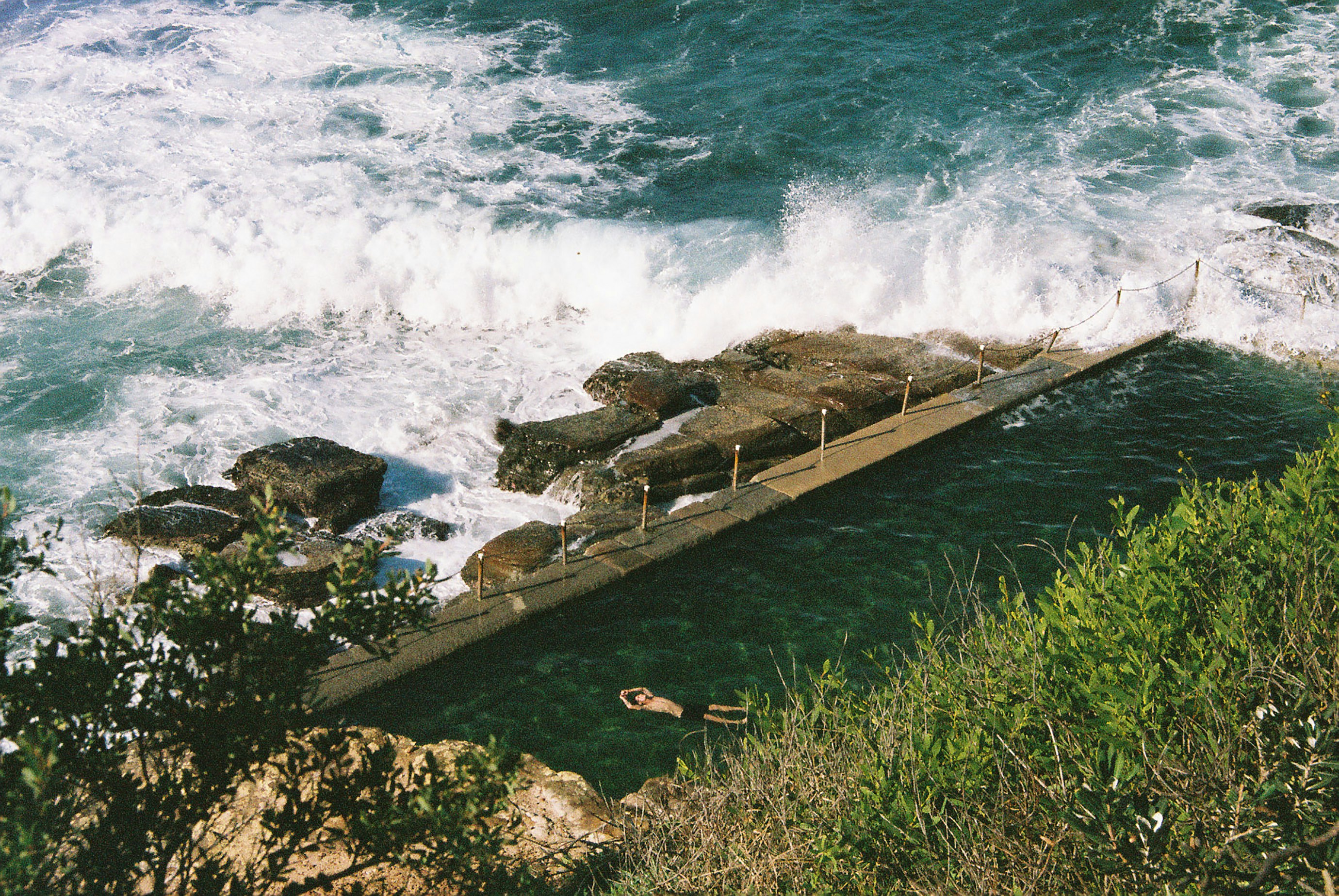 Rectangular pool hugs the shoreline as waves crash over rocks beyond a railing, with dense greenery in the foreground framing the scene.