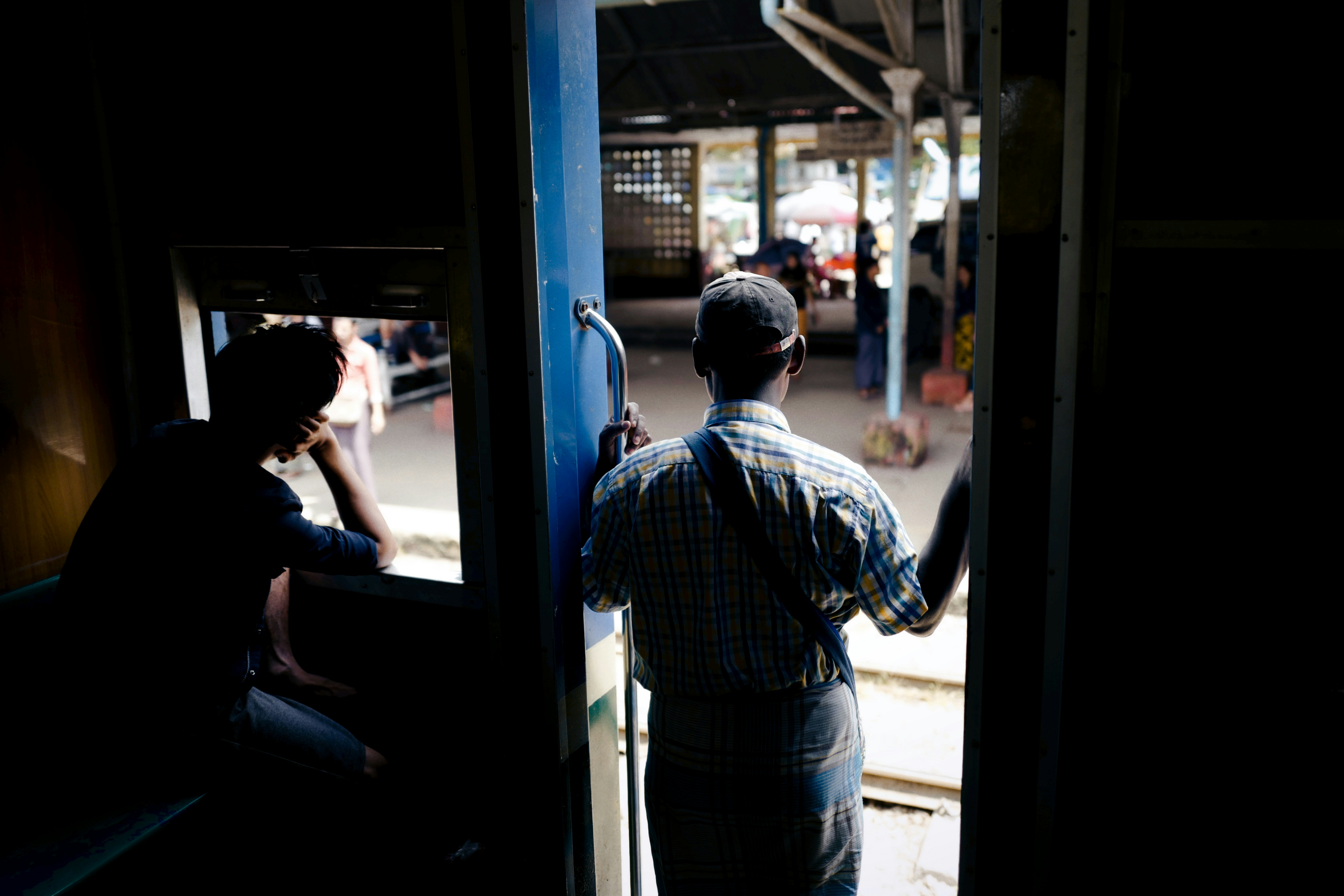 A man stands at the entrance of a train, gazing out into the vibrant market beyond, while another passenger sits in contemplation. 