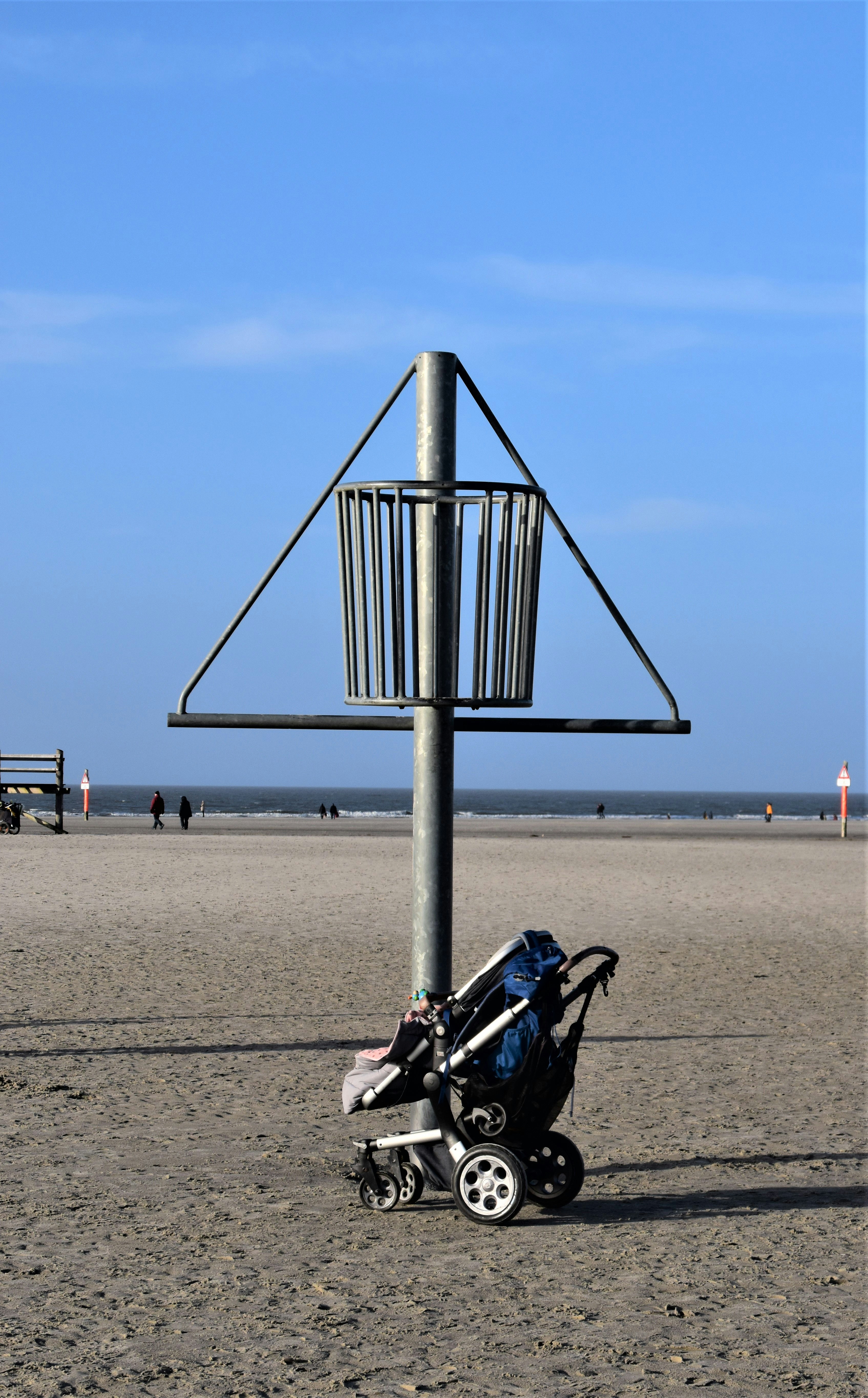 A beach scene featuring a unique metal wastebasket structure alongside a parked stroller, highlighting the blend of utility and leisure in outdoor spaces.