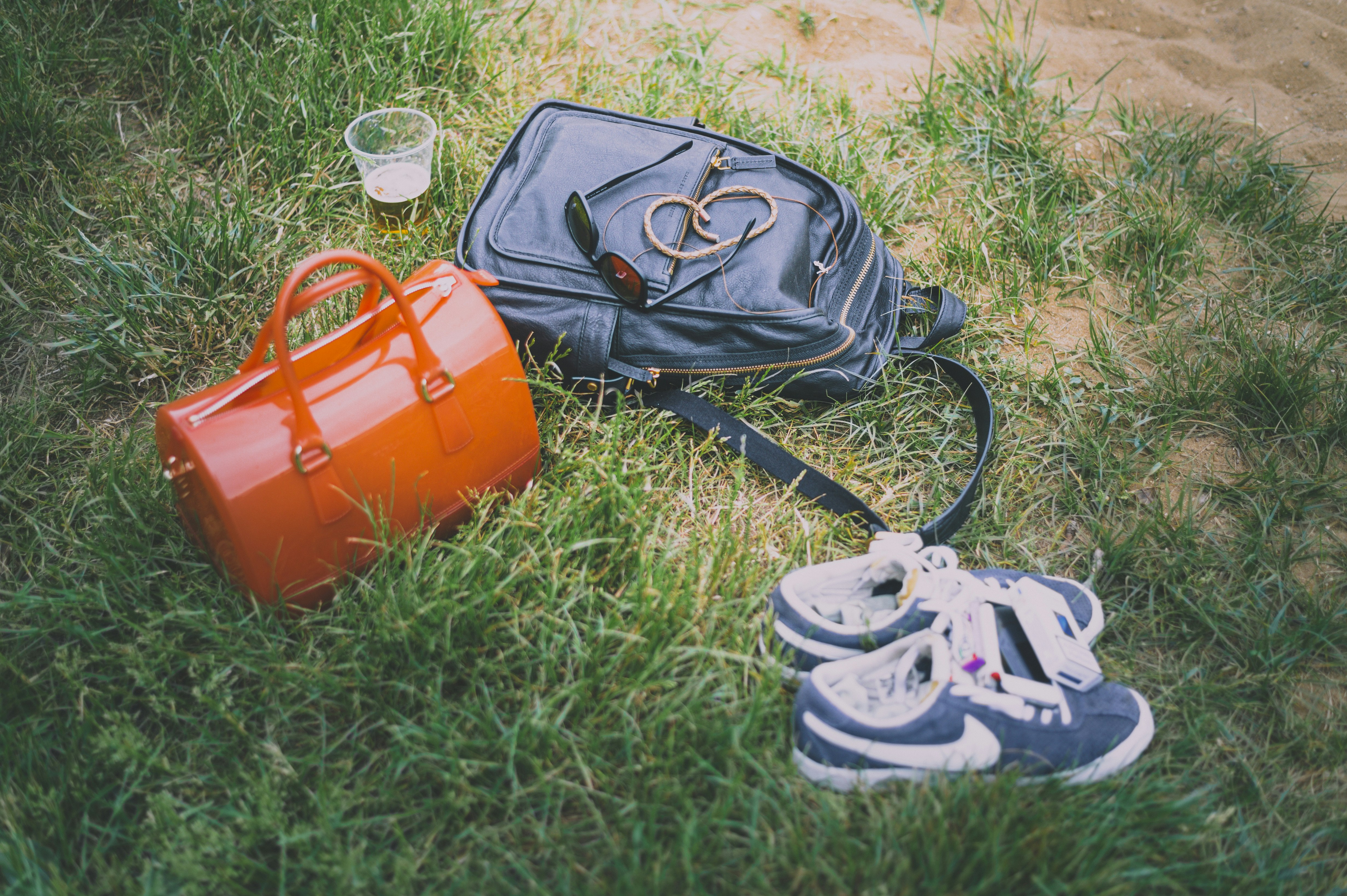 An orange handbag, a black backpack, sunglasses, and sneakers arranged on lush green grass, evoking a relaxed outdoor vibe.